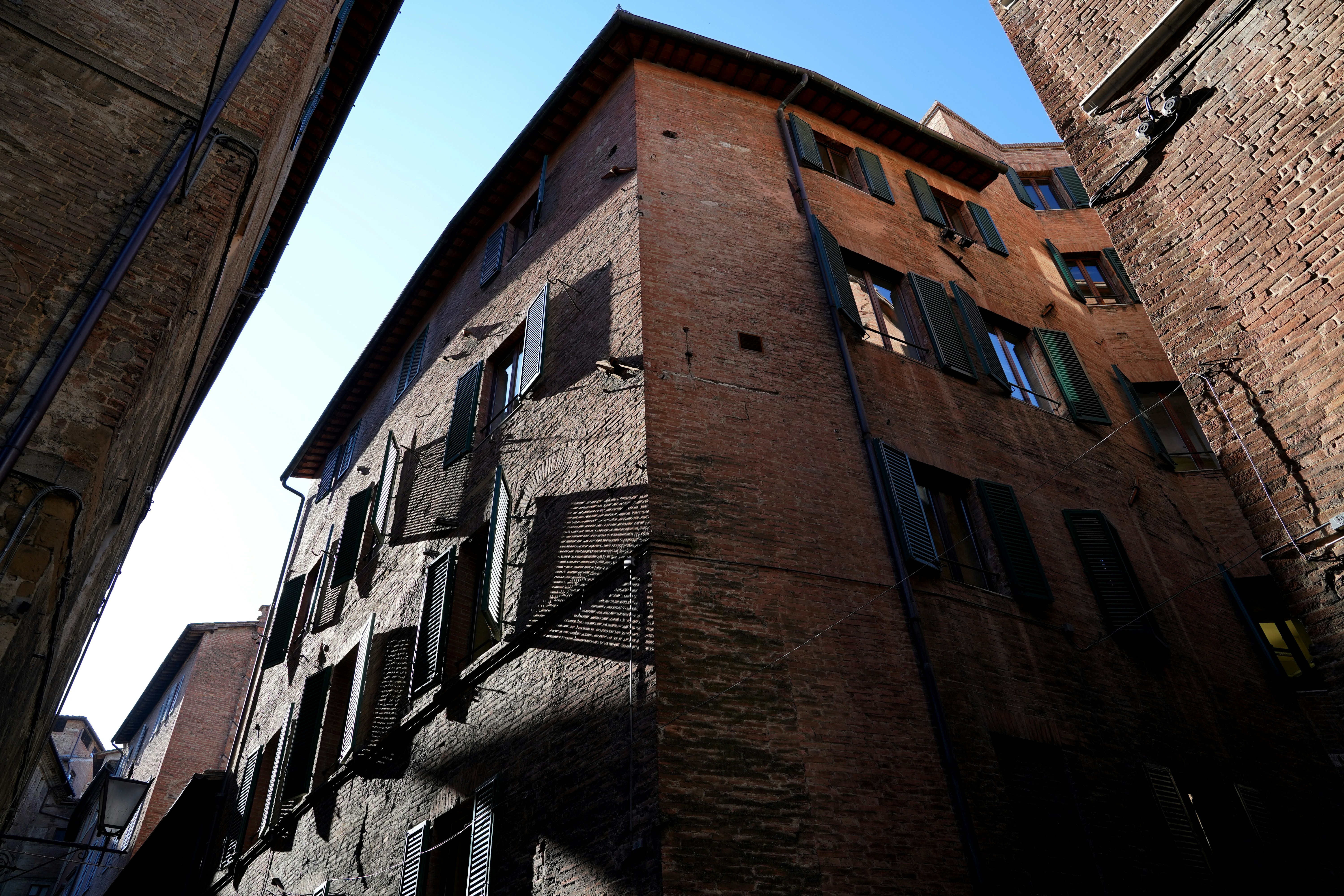 Tall brick building with wooden shutters casts dramatic shadows under a clear blue sky.