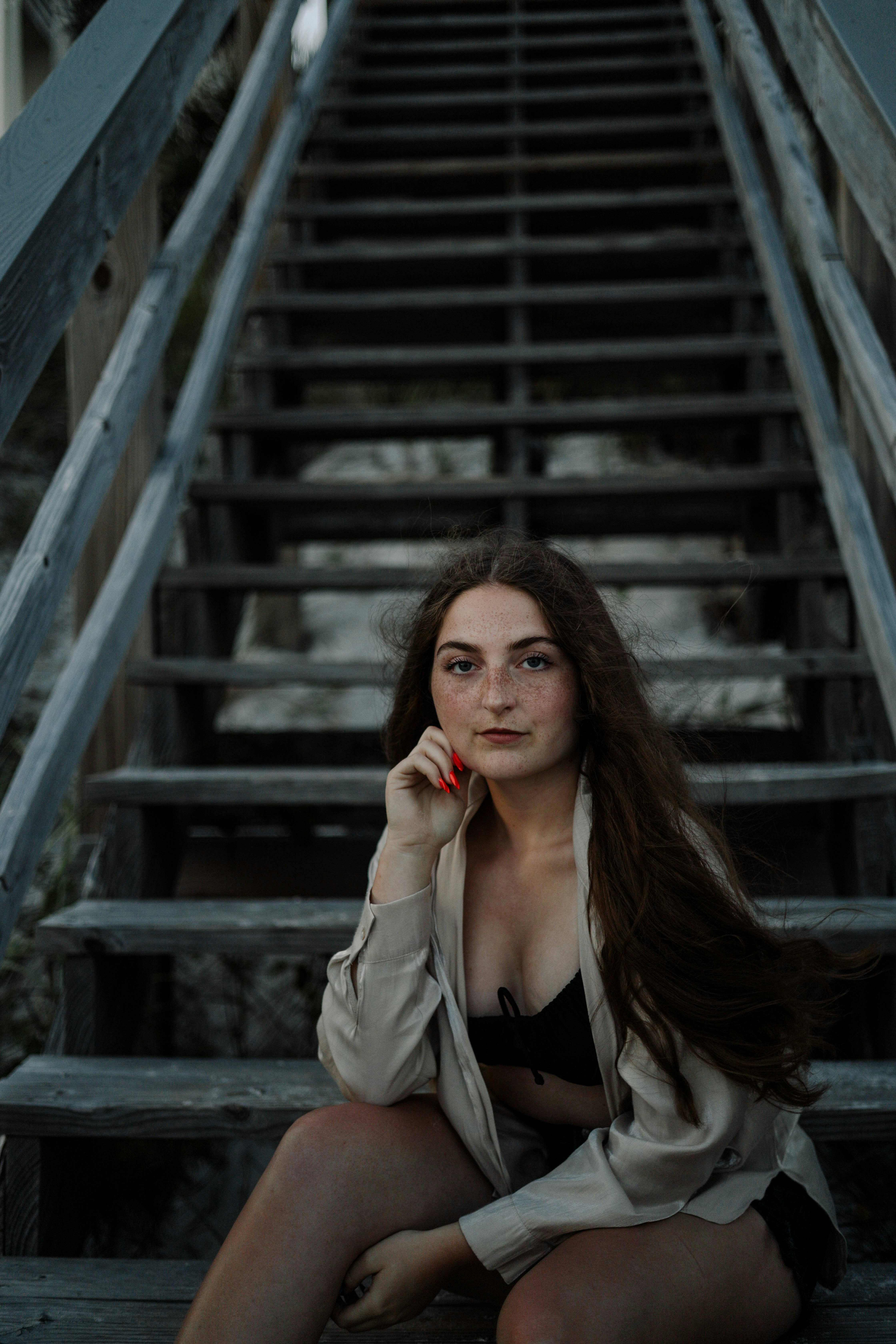 Young woman seated on wooden stairs, exuding confidence with a casual pose and striking red nails.