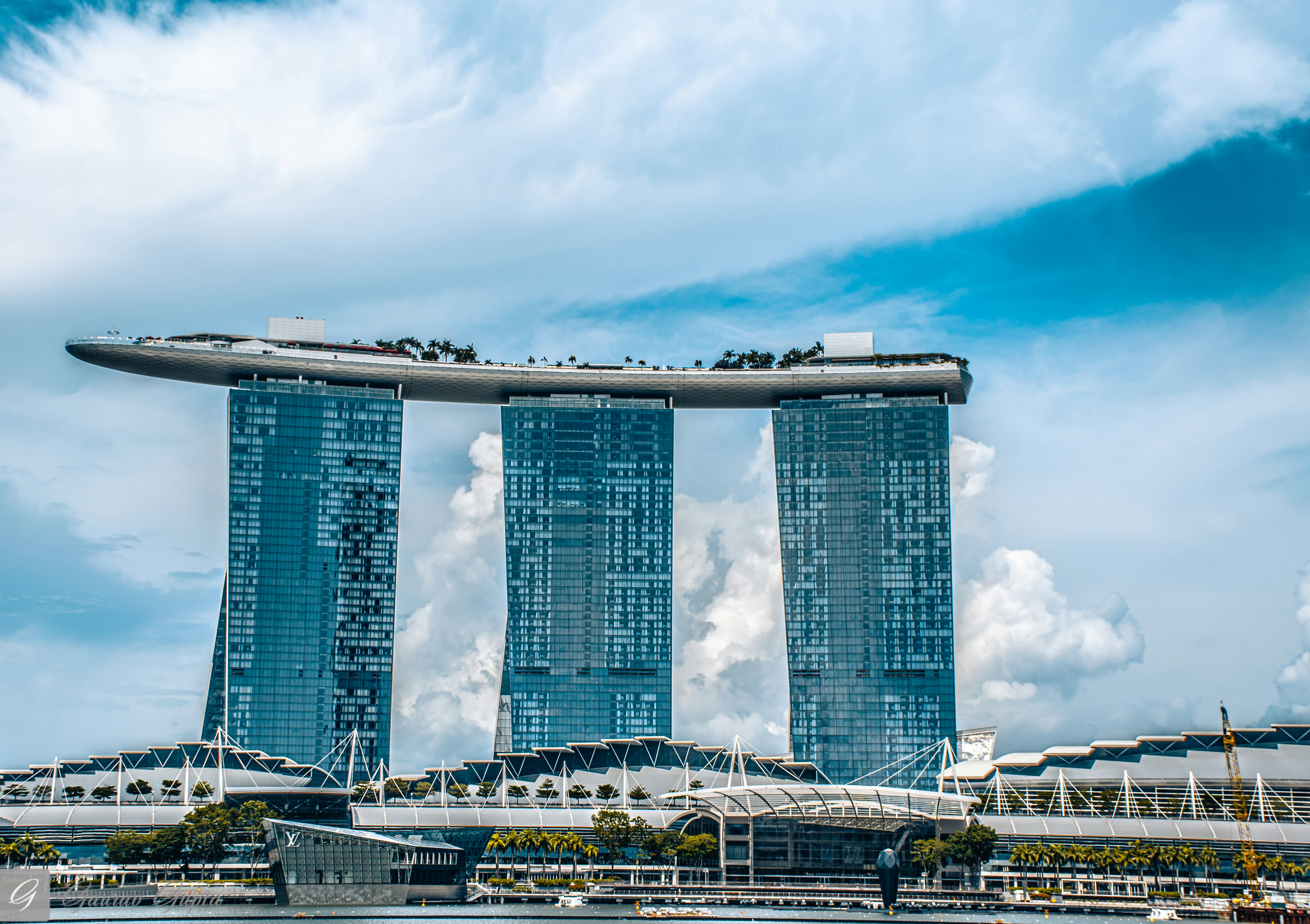 Iconic Marina Bay Sands towers with a unique boat-shaped structure atop, set against a vibrant blue sky with scattered clouds.