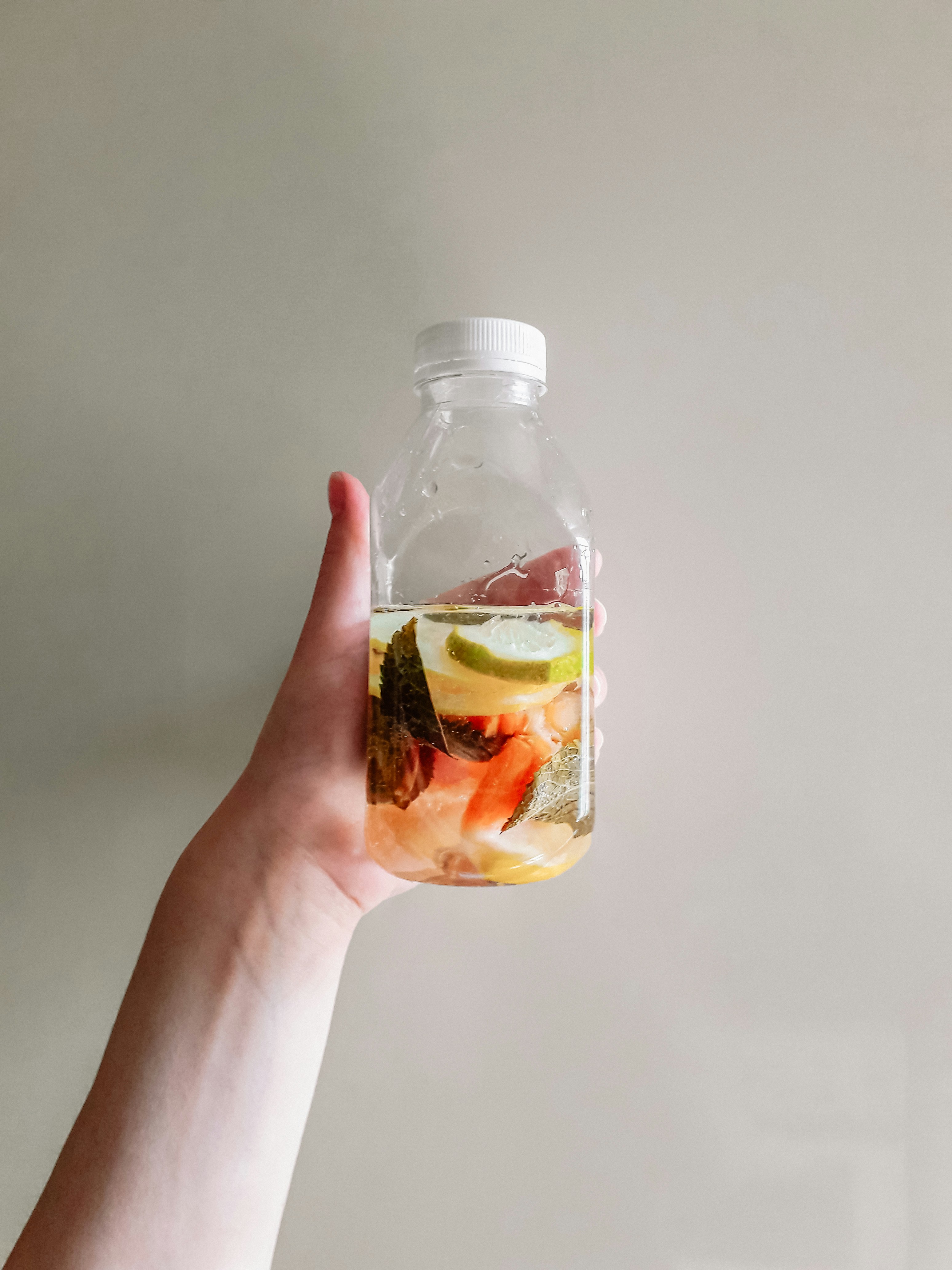 A hand holding a transparent bottle filled with infused water featuring slices of lime, strawberries, and mint leaves against a neutral background.