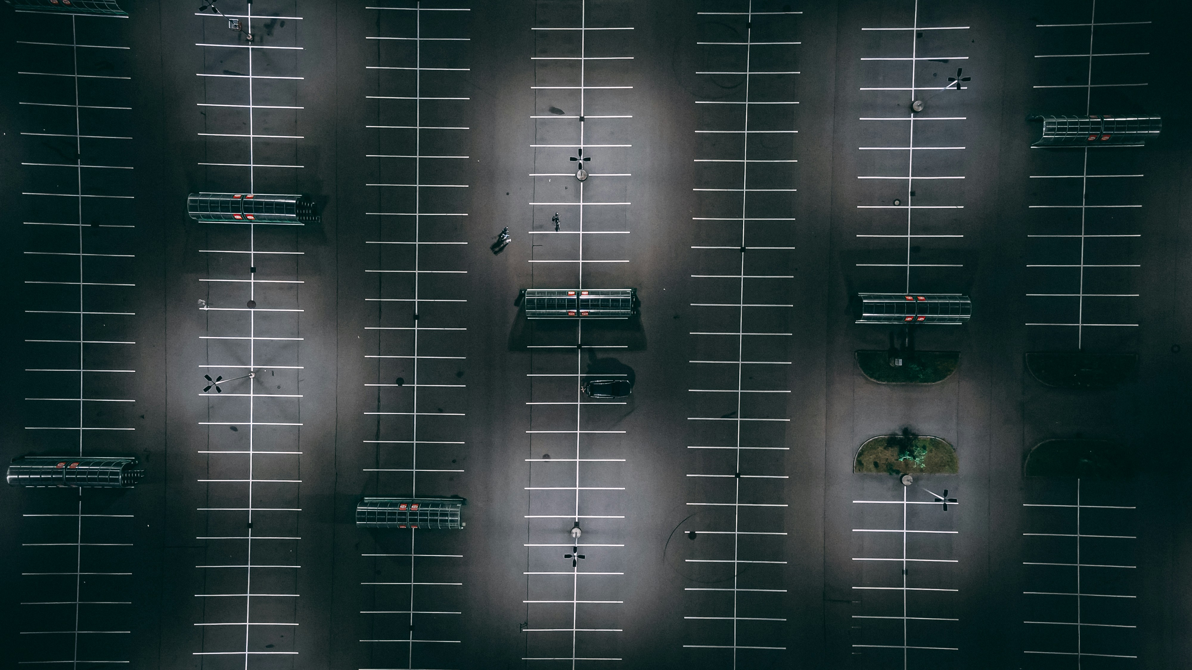 Aerial view of an expansive parking lot, showcasing a grid of empty spaces with a few scattered vehicles and pedestrians. The stark lines create a sense of isolation.