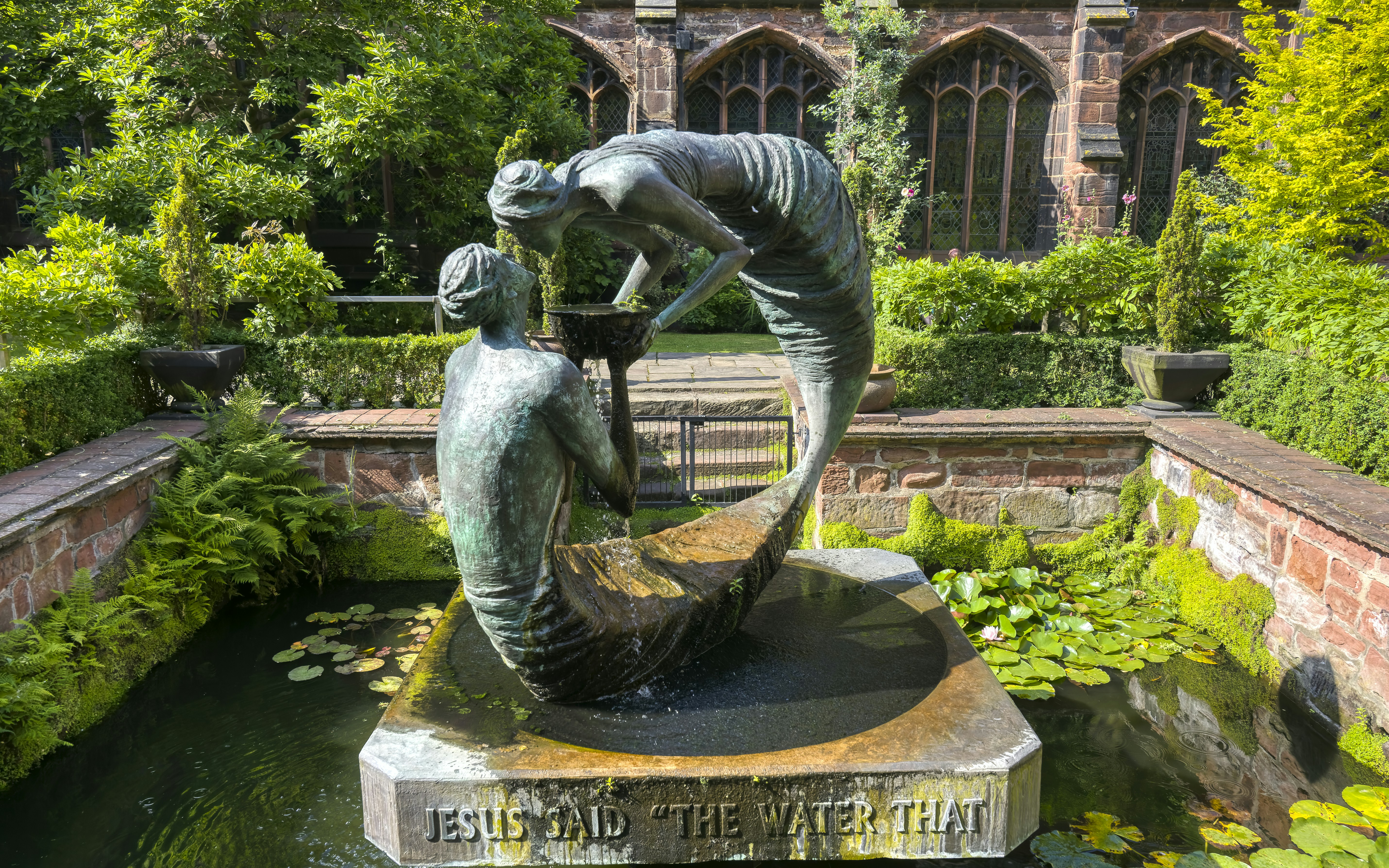 Bronze sculpture depicting a figure leaning over another, surrounded by lush greenery and water features in a tranquil garden setting.