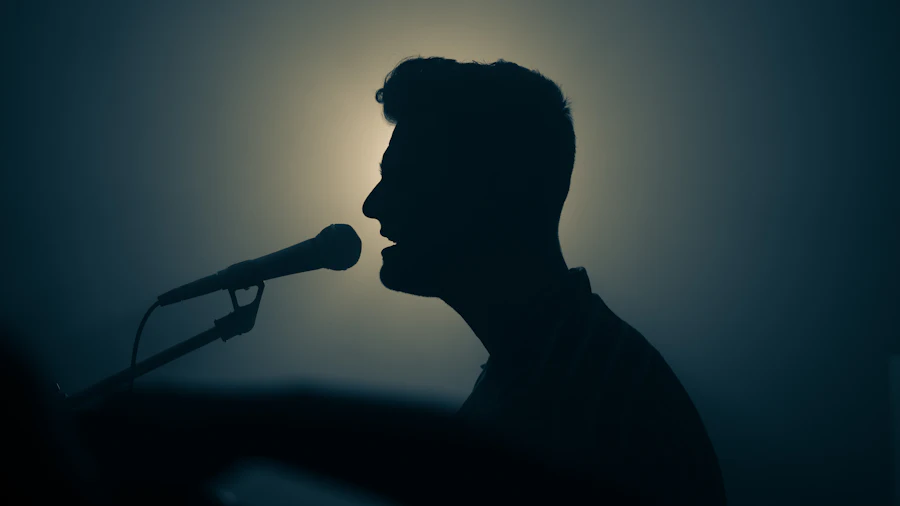 Side-profile silhouette of a singer leaning into a microphone with a soft backlight glowing behind the head. Dark stage foreground and moody teal shadows frame the scene.