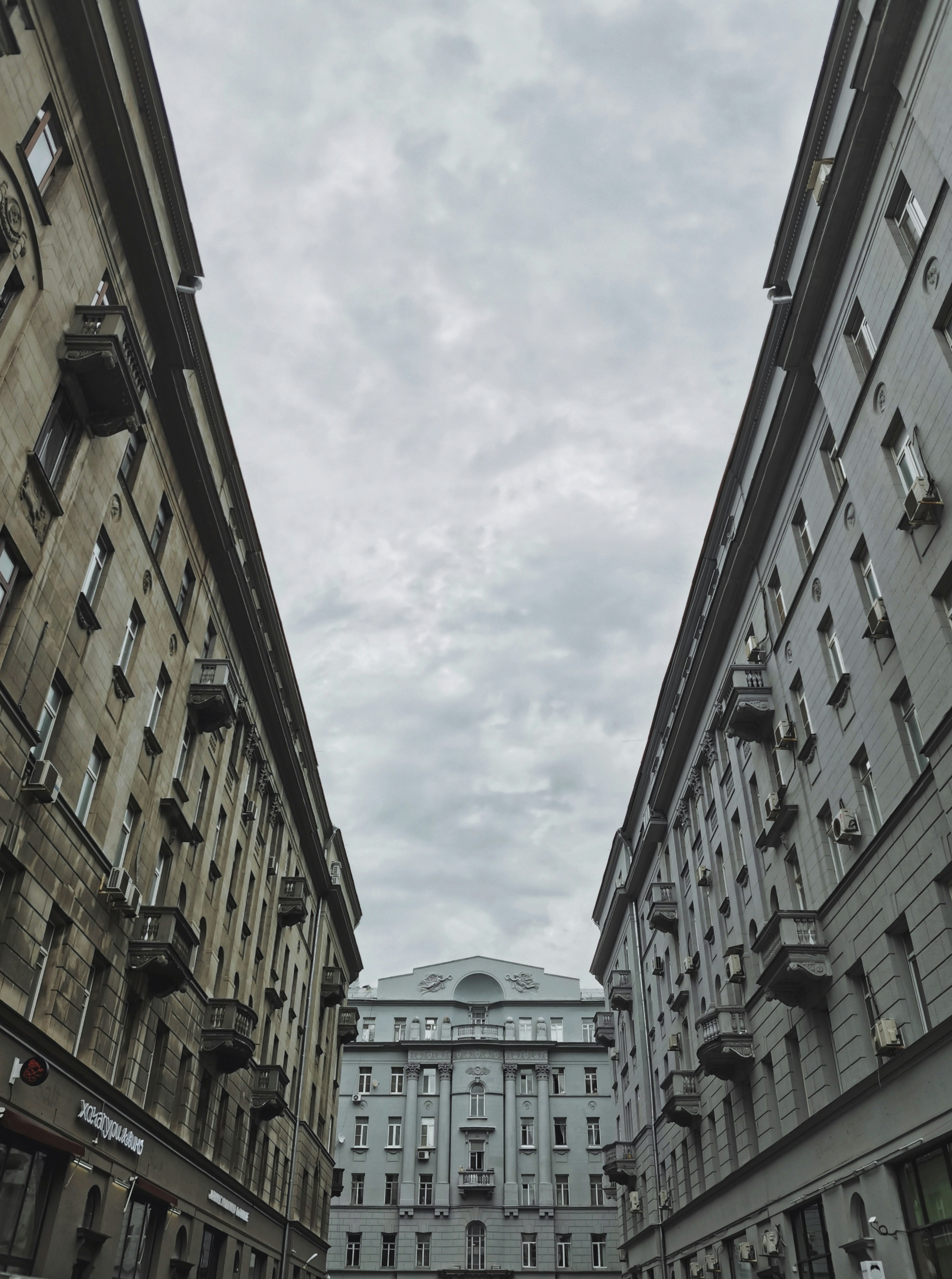Tall, closely spaced buildings frame a narrow street that converges toward a distant, ornate façade under a cloudy sky.
