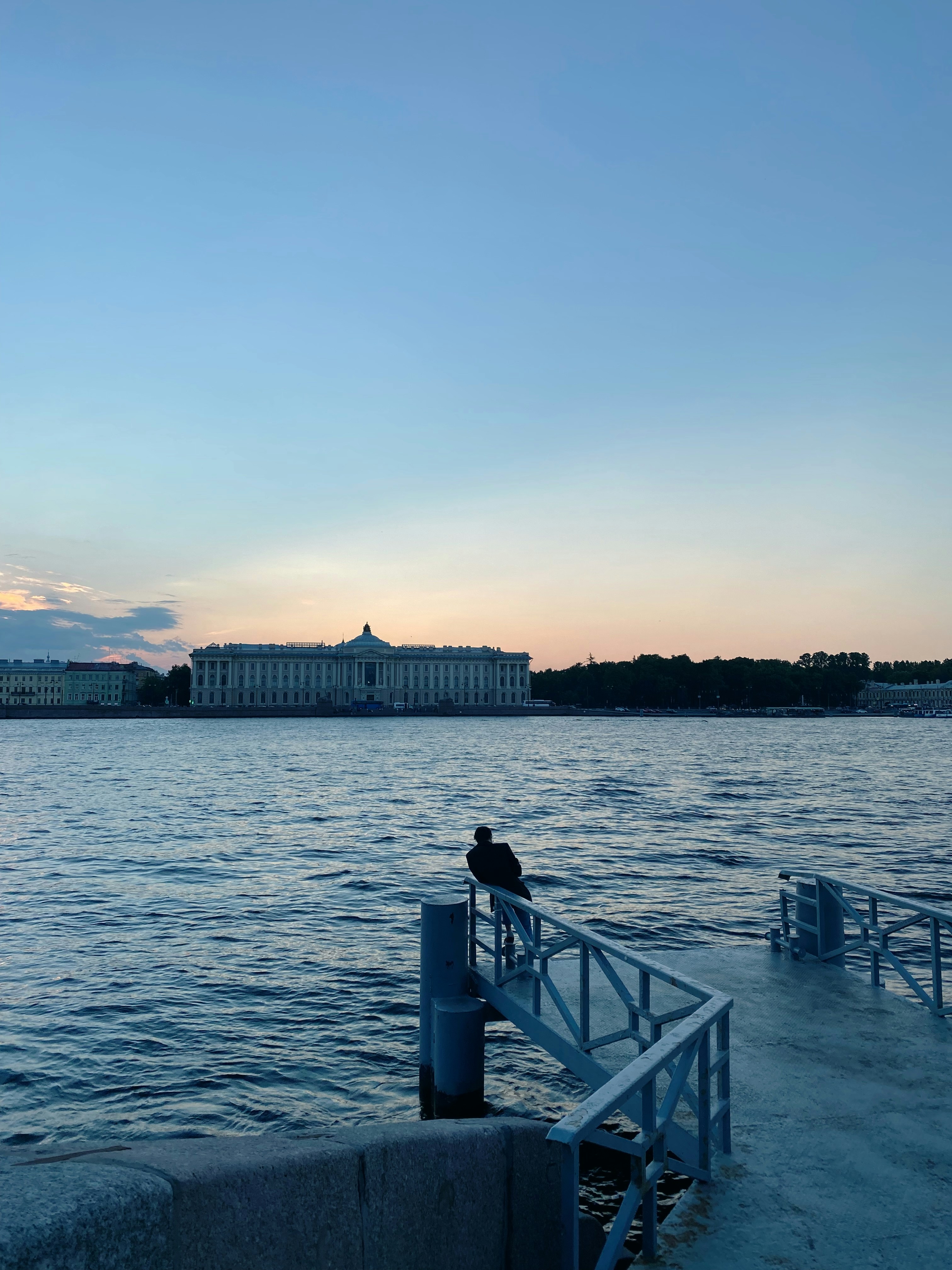 A lone figure sitting on a dock, gazing at the tranquil waters under a twilight sky, with an impressive building in the background.