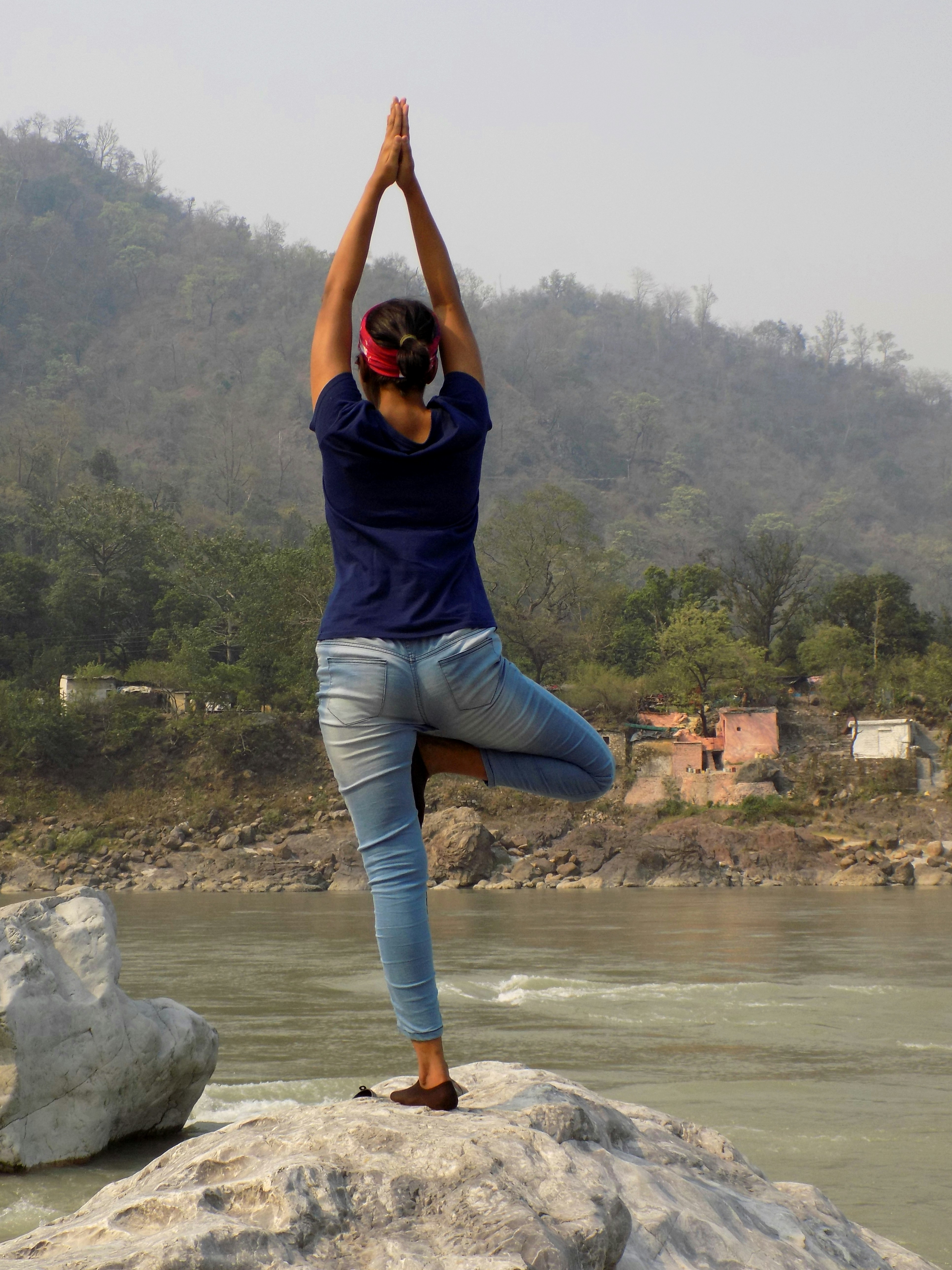 A woman practicing yoga in a tree pose on a rock by a flowing river, surrounded by lush greenery and distant homes.