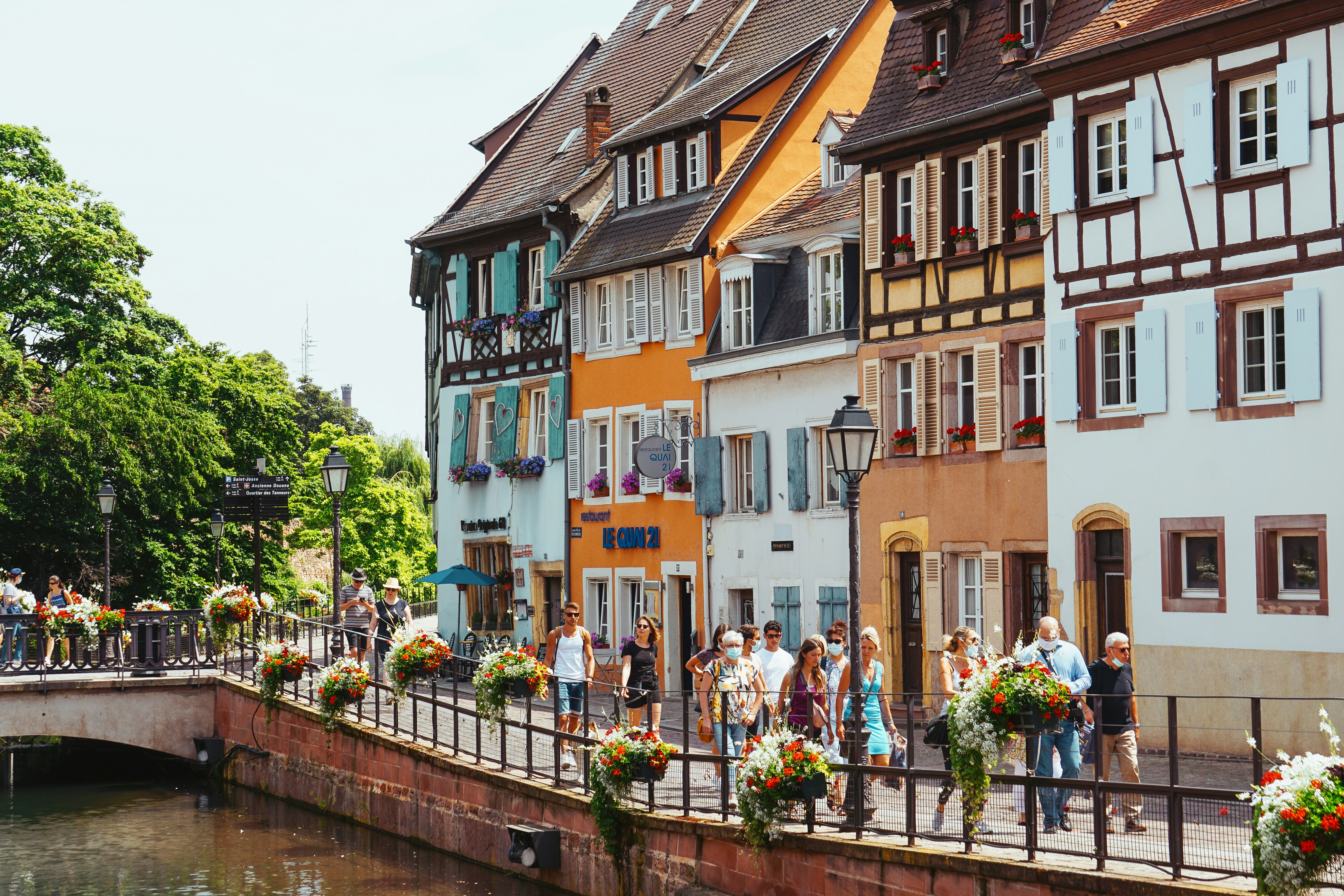 Vibrant, half-timbered buildings line a canal in a charming European town, with people walking along the flower-adorned path.