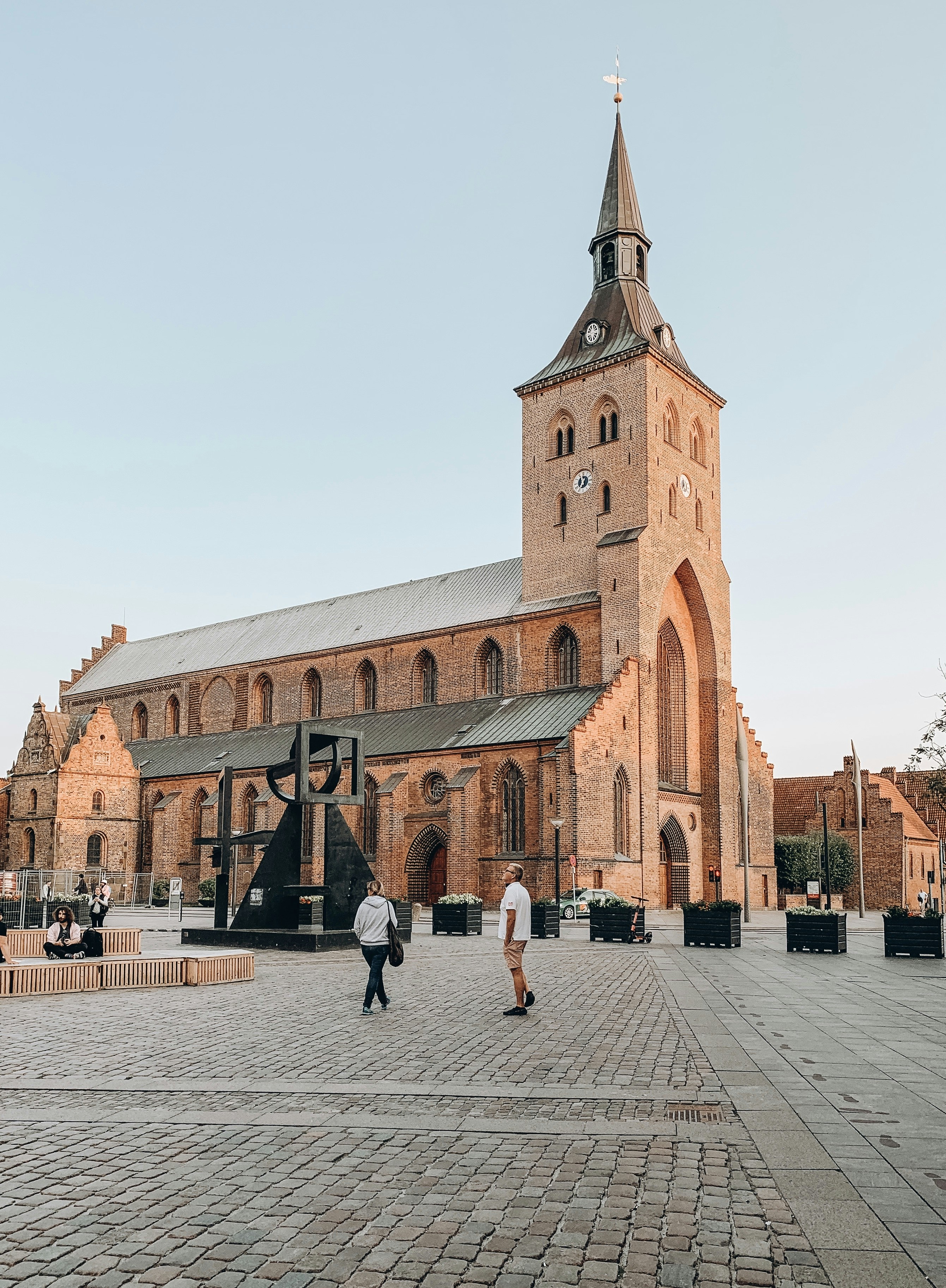 Historic cathedral with a tall spire under a clear evening sky, two people walking across a cobblestone plaza.