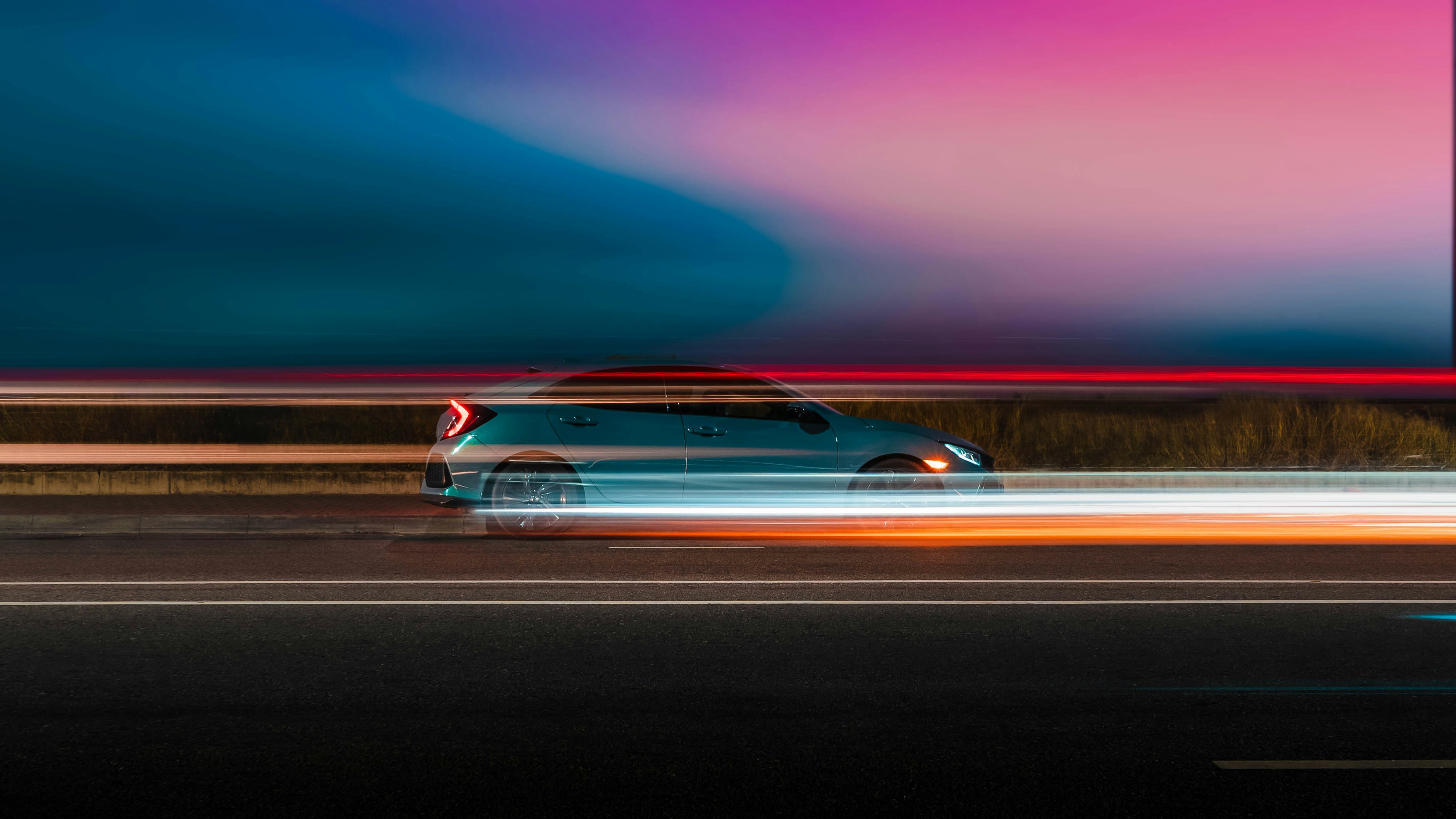 A sleek car glides along a highway, captured in motion against a vibrant twilight sky with streaks of light from passing vehicles.