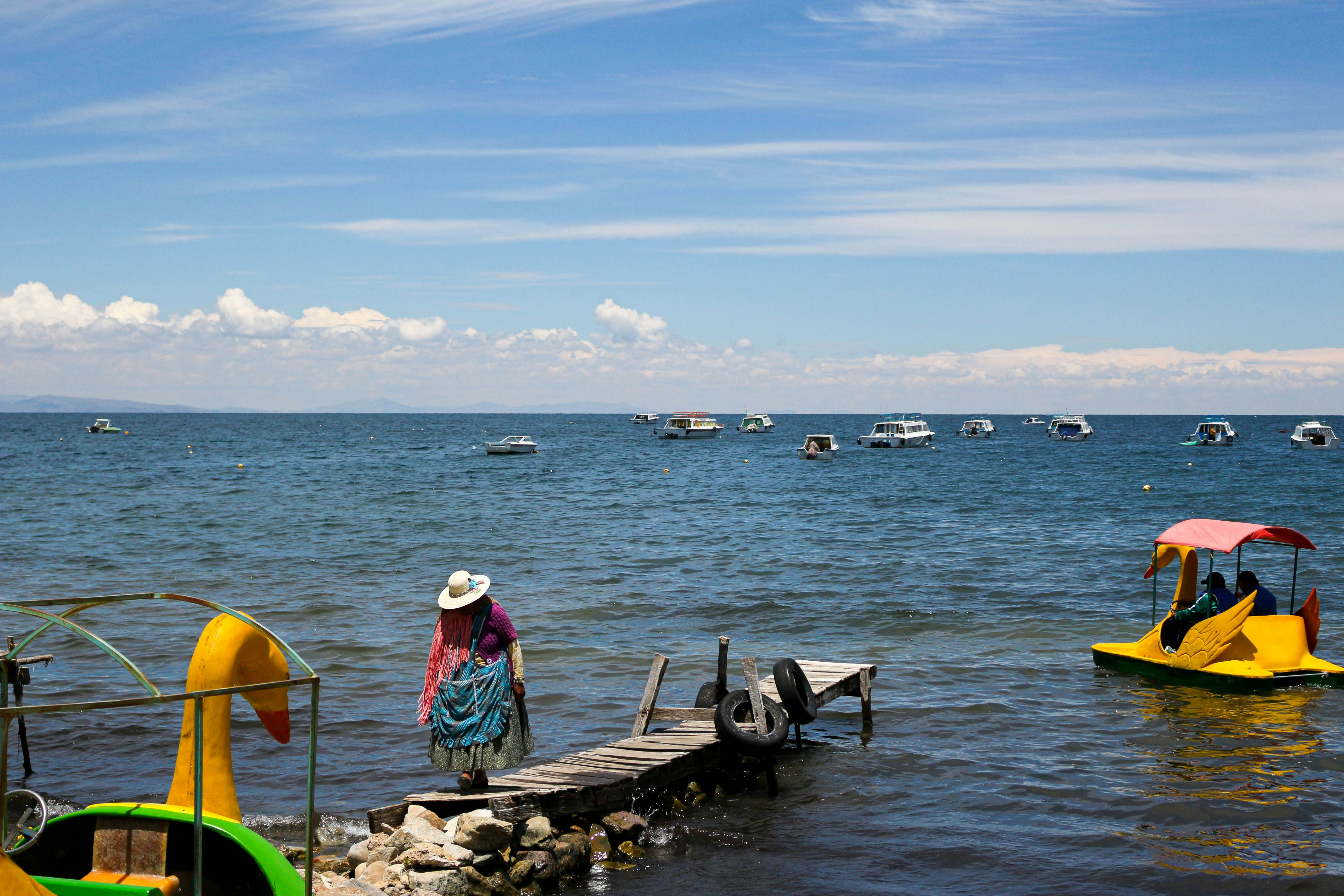 Woman in traditional attire stands on a wooden pier overlooking a lake with boats in the distance.