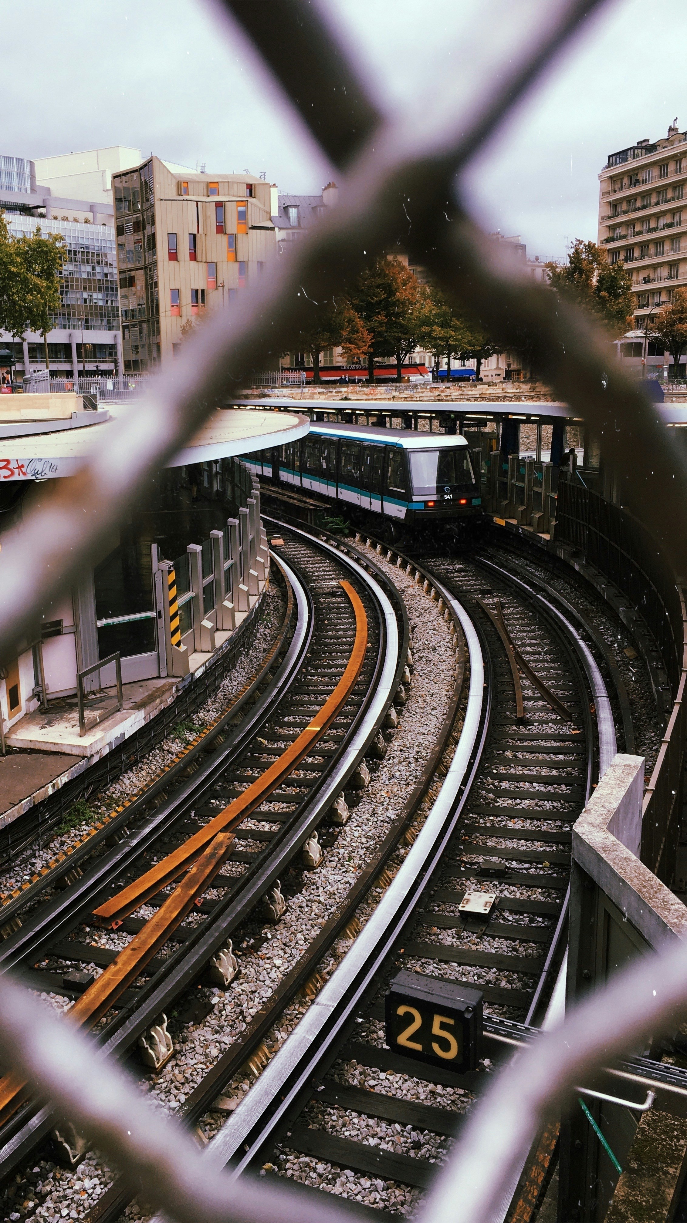 Curving train tracks leading into a station, viewed through a chain-link fence, showcasing the interplay of urban architecture and transportation.