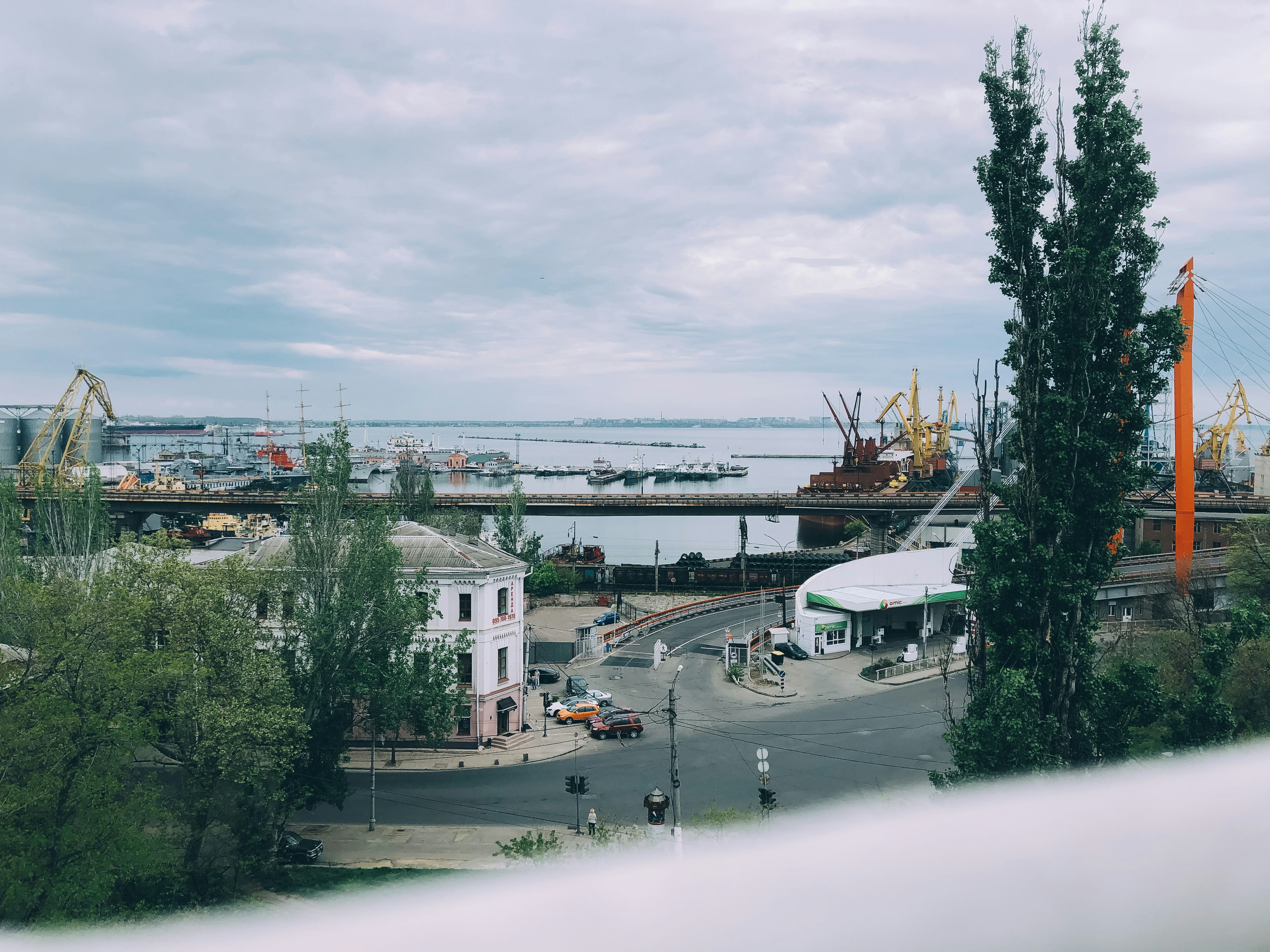 Urban scene with industrial harbor and overcast sky, framed by tall trees and a curved road.