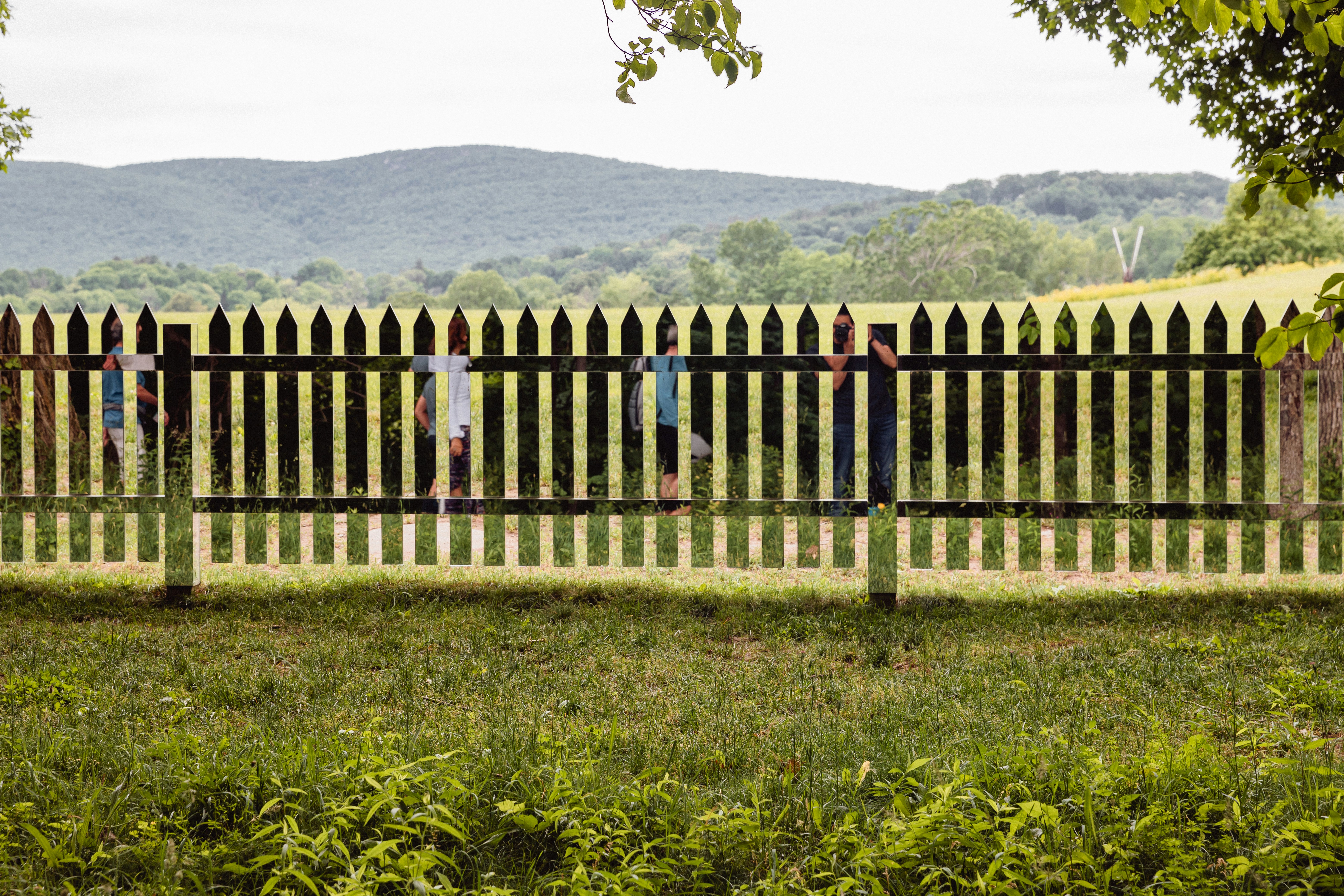 People walking along a path framed by a white picket fence, with rolling hills in the background. The scene captures a serene rural landscape.