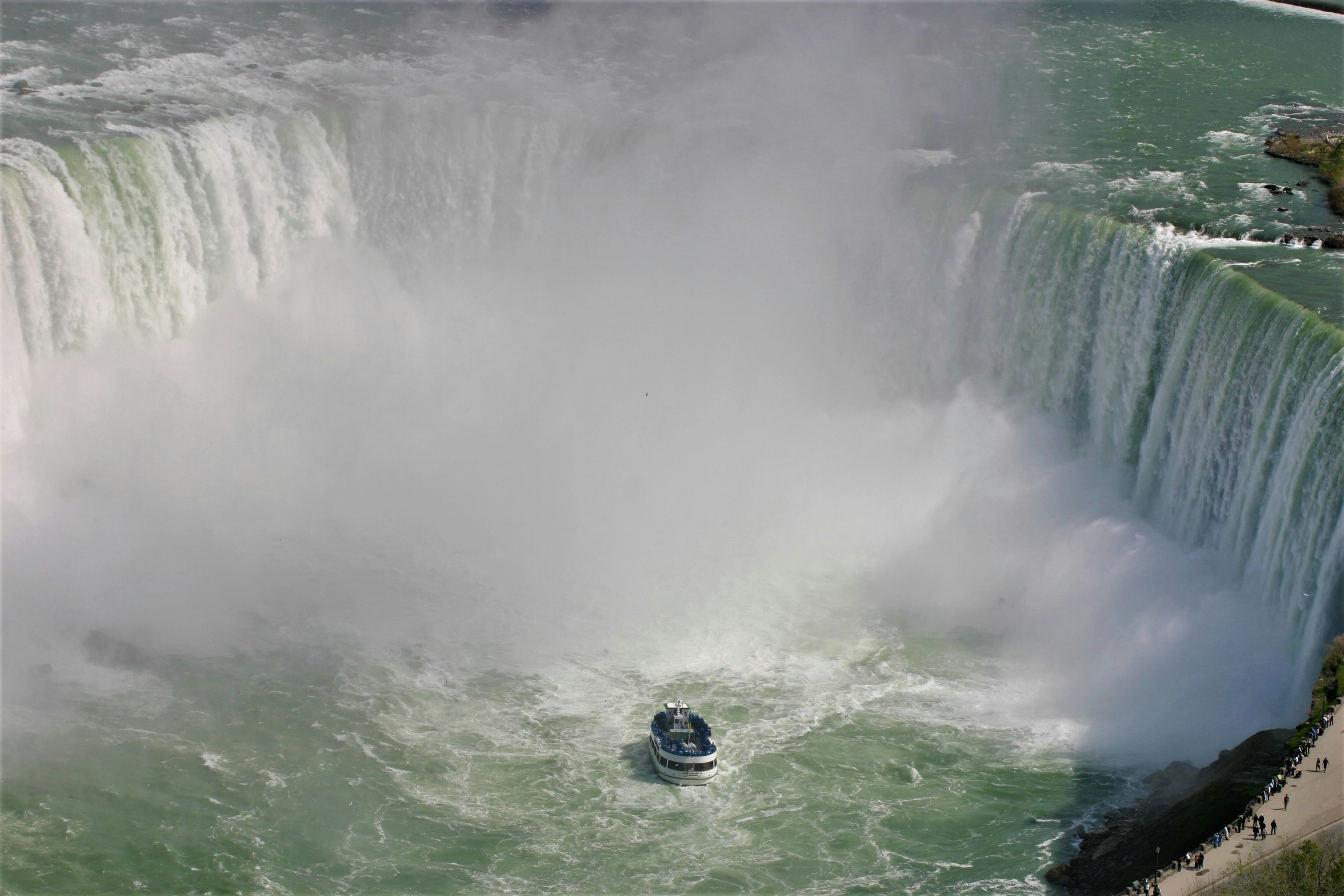 Tourist boat navigating the churning waters near the base of a massive waterfall, surrounded by mist and cascading water.