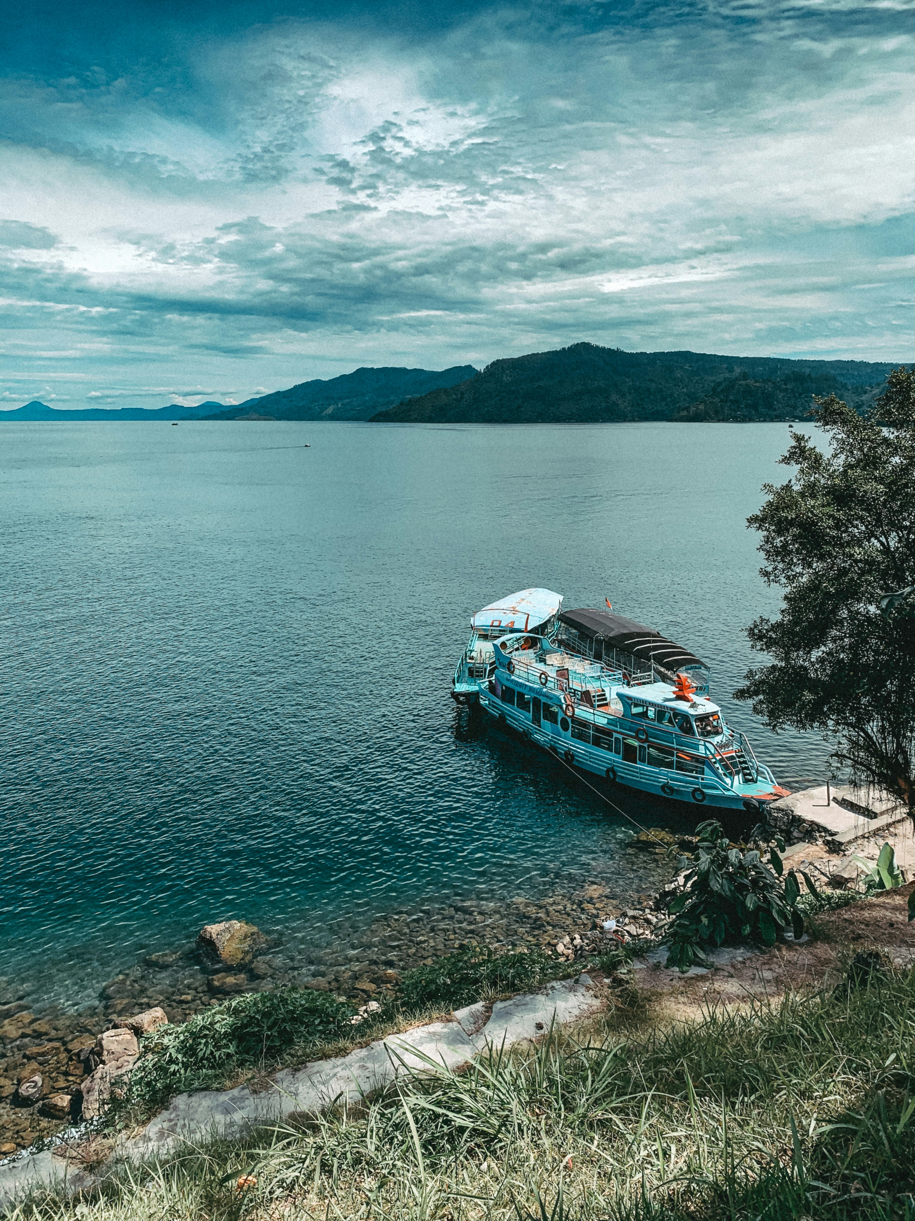 A colorful boat docked near a rocky shoreline, surrounded by lush greenery and calm waters under a cloudy sky.