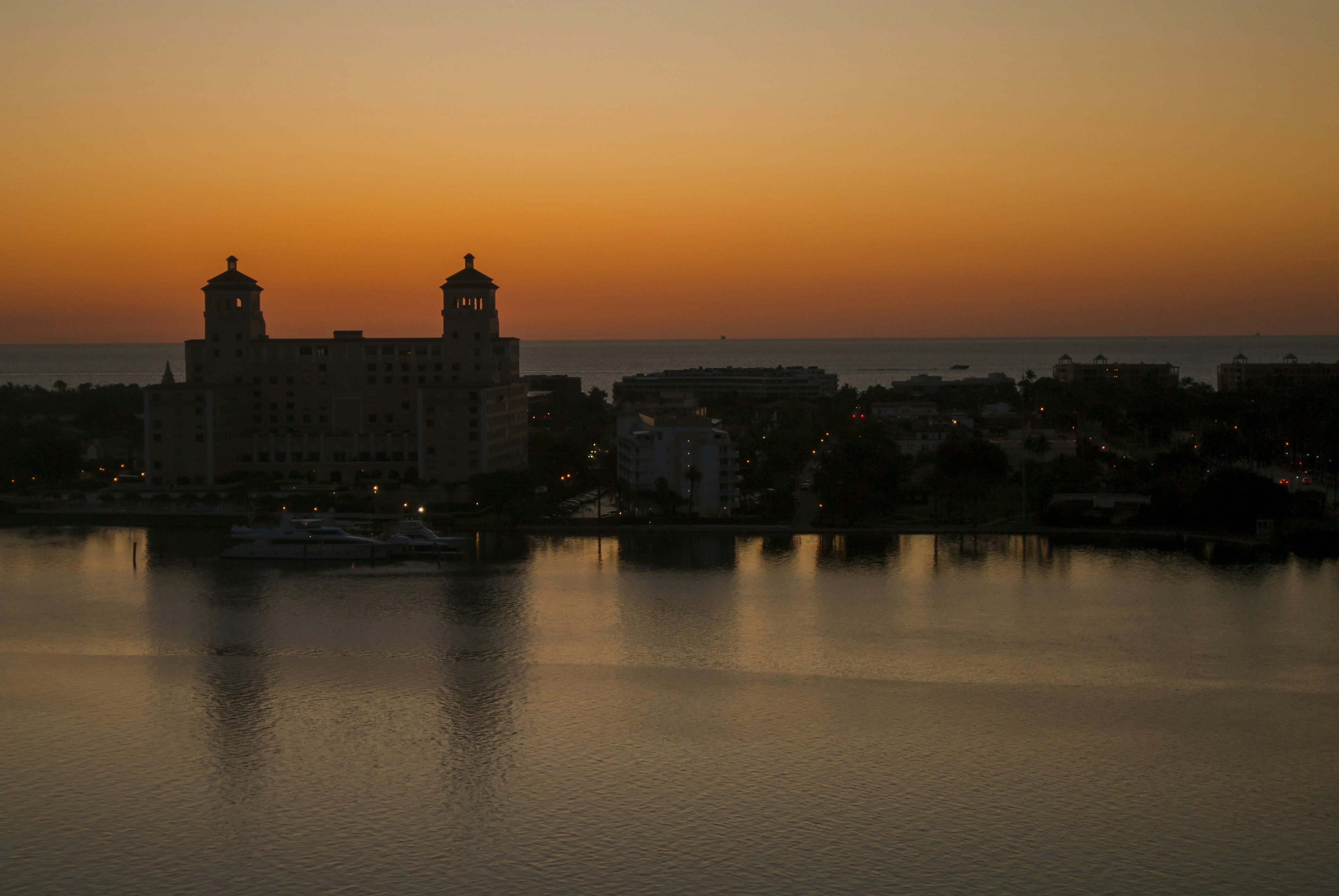 Silhouetted buildings against a gradient sunset, casting reflections on calm waters. The scene captures the tranquil essence of twilight by the coast.