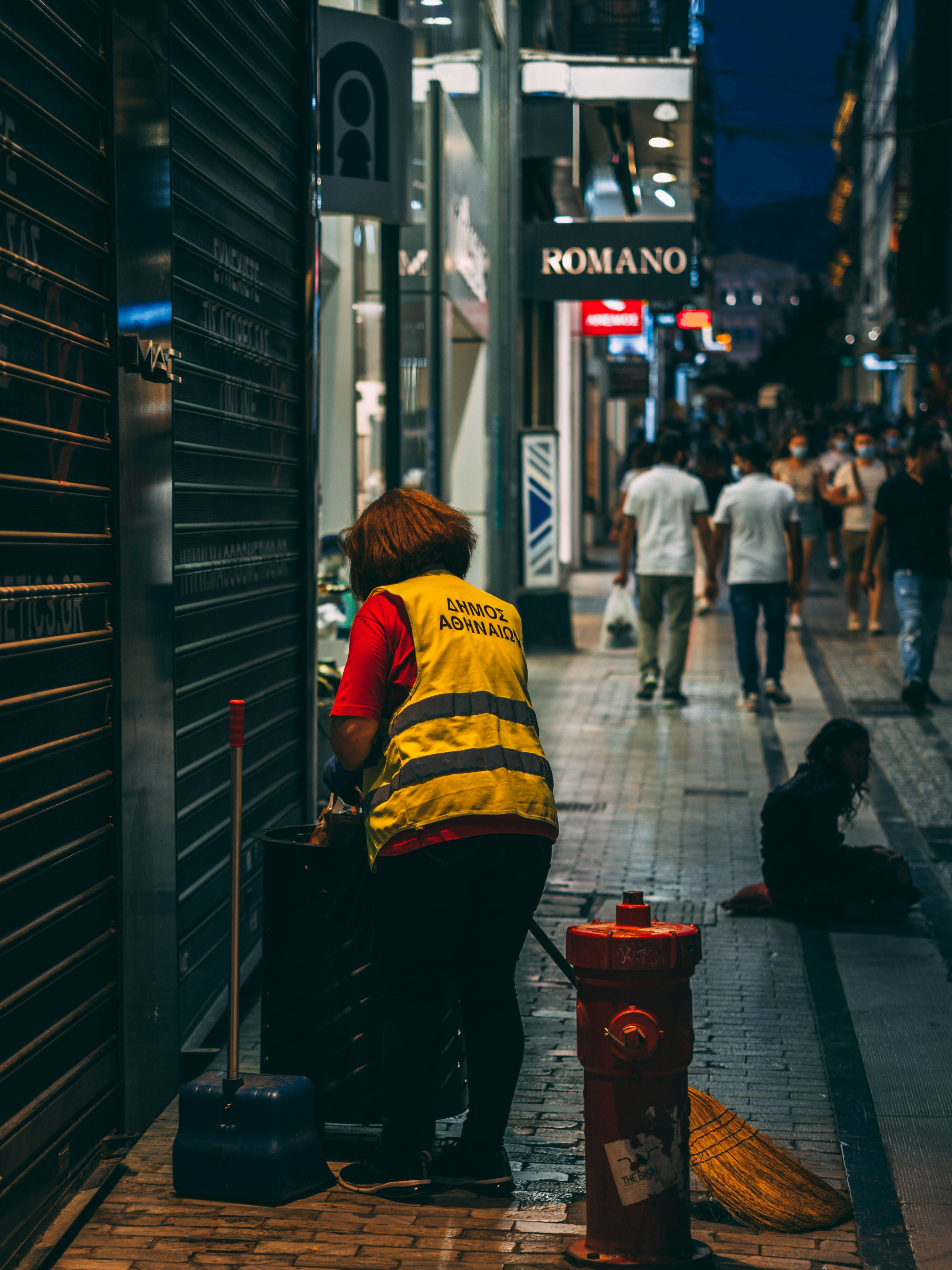 A worker in a reflective vest tending to tasks on a bustling street at night, surrounded by pedestrians and illuminated storefronts.