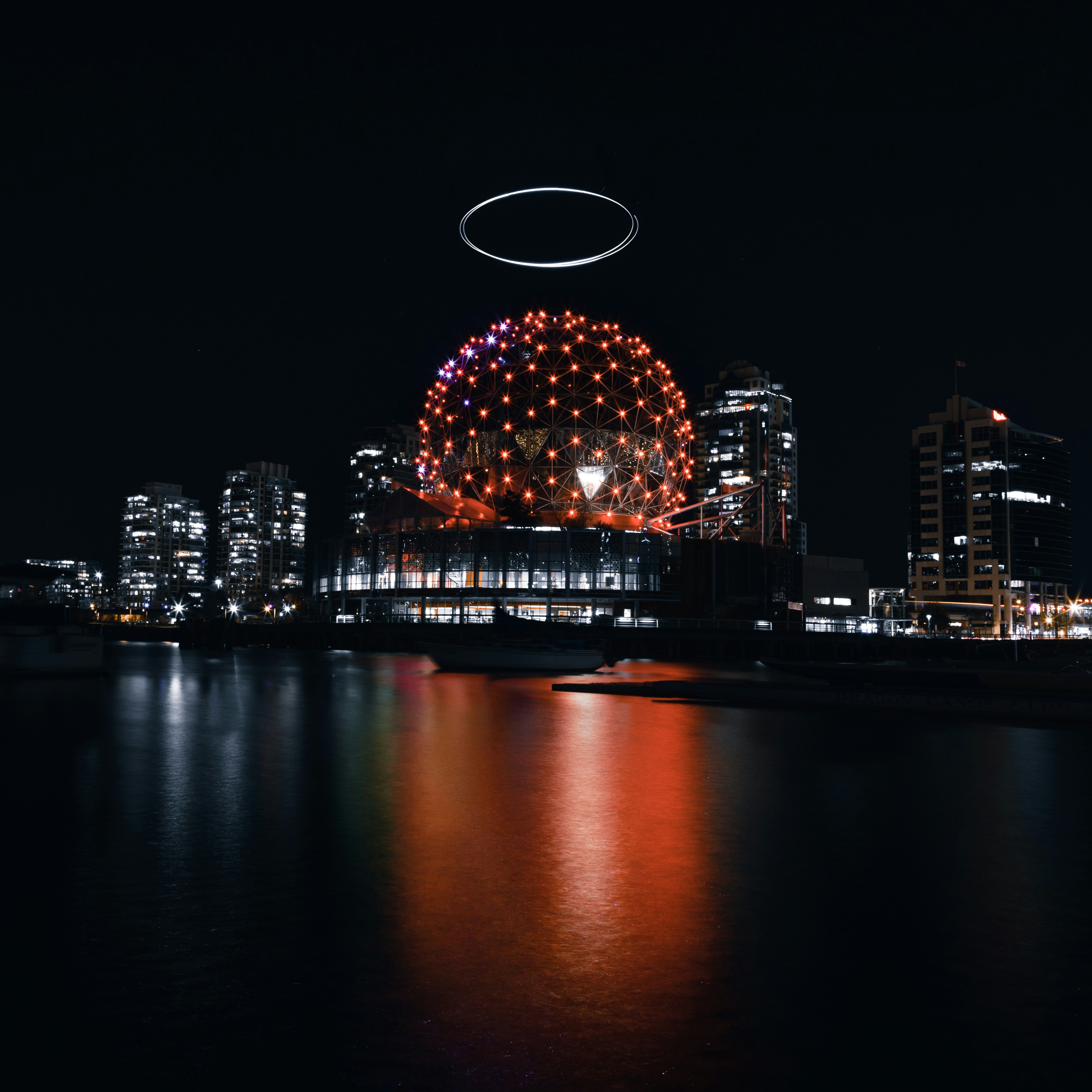 Vibrant illuminated dome reflecting on water, surrounded by city skyline at night.
