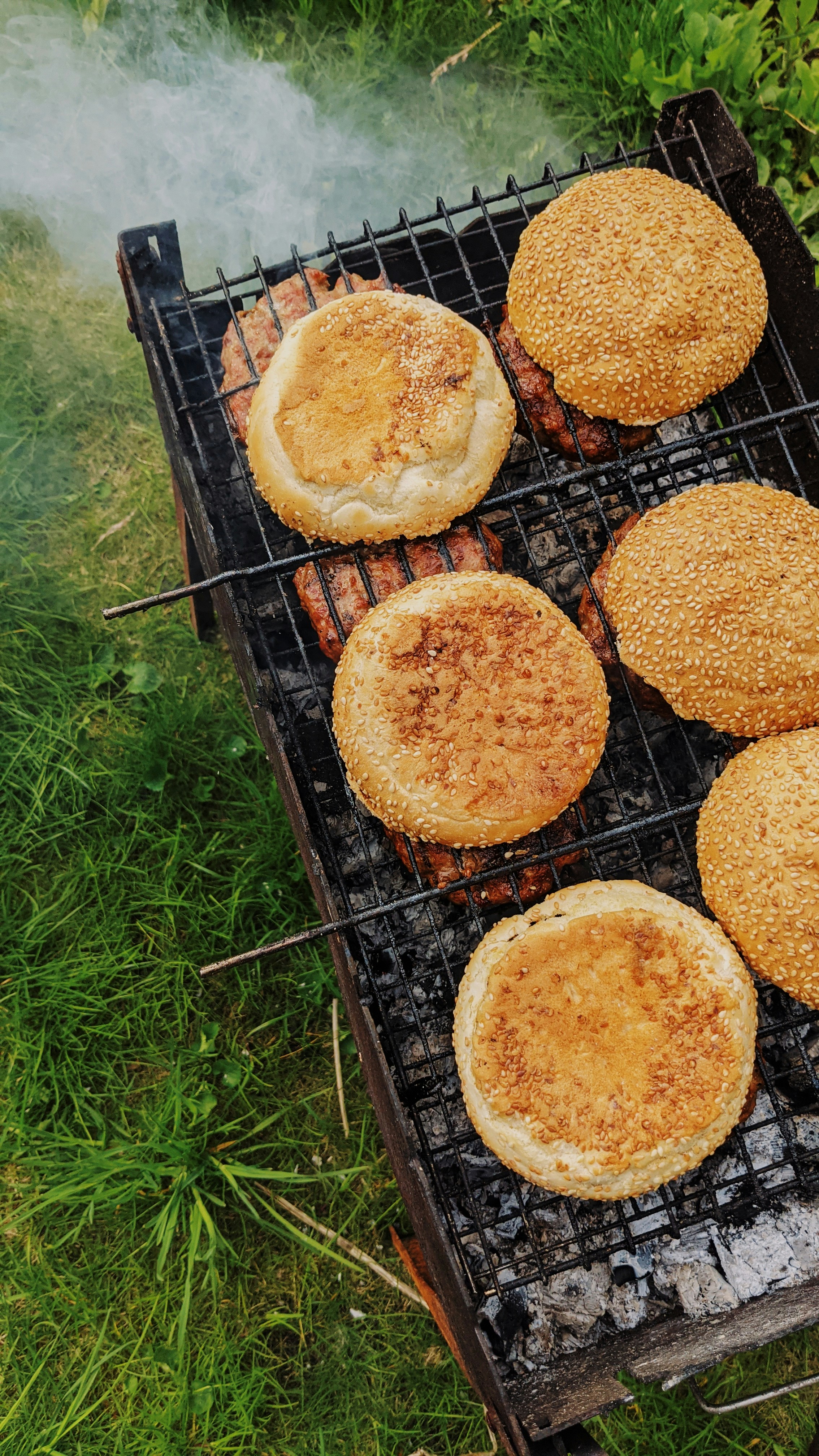 Sesame seed burger buns toasting on a charcoal grill, surrounded by wisps of smoke. The scene captures the essence of outdoor cooking.