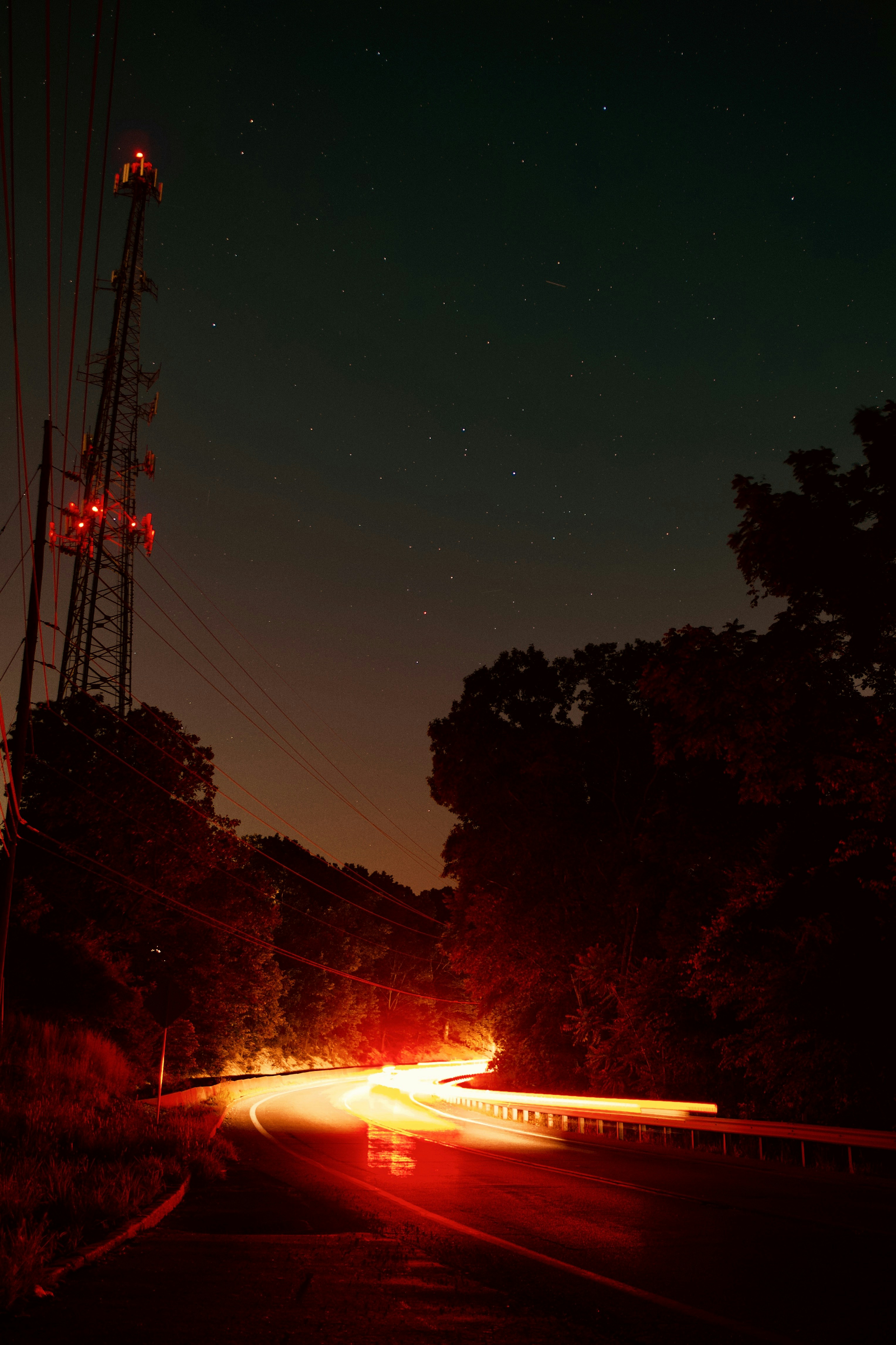 Long exposure capturing the winding road illuminated by car lights under a starry sky, with a telecommunications tower in the foreground.
