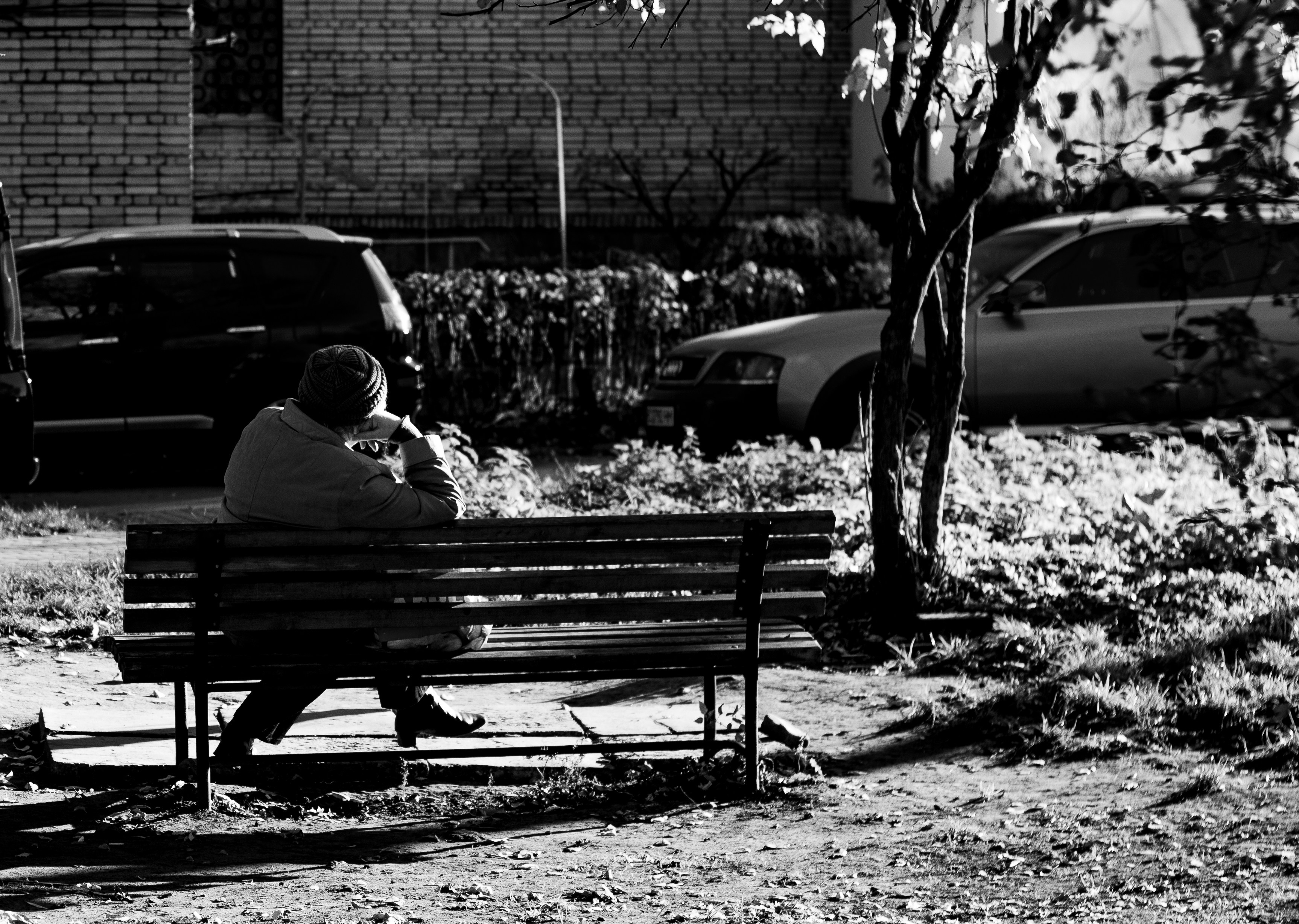 A solitary figure sits on a bench, immersed in thought, surrounded by the quiet bustle of an urban park. The scene is captured in high contrast black and white, enhancing the mood.