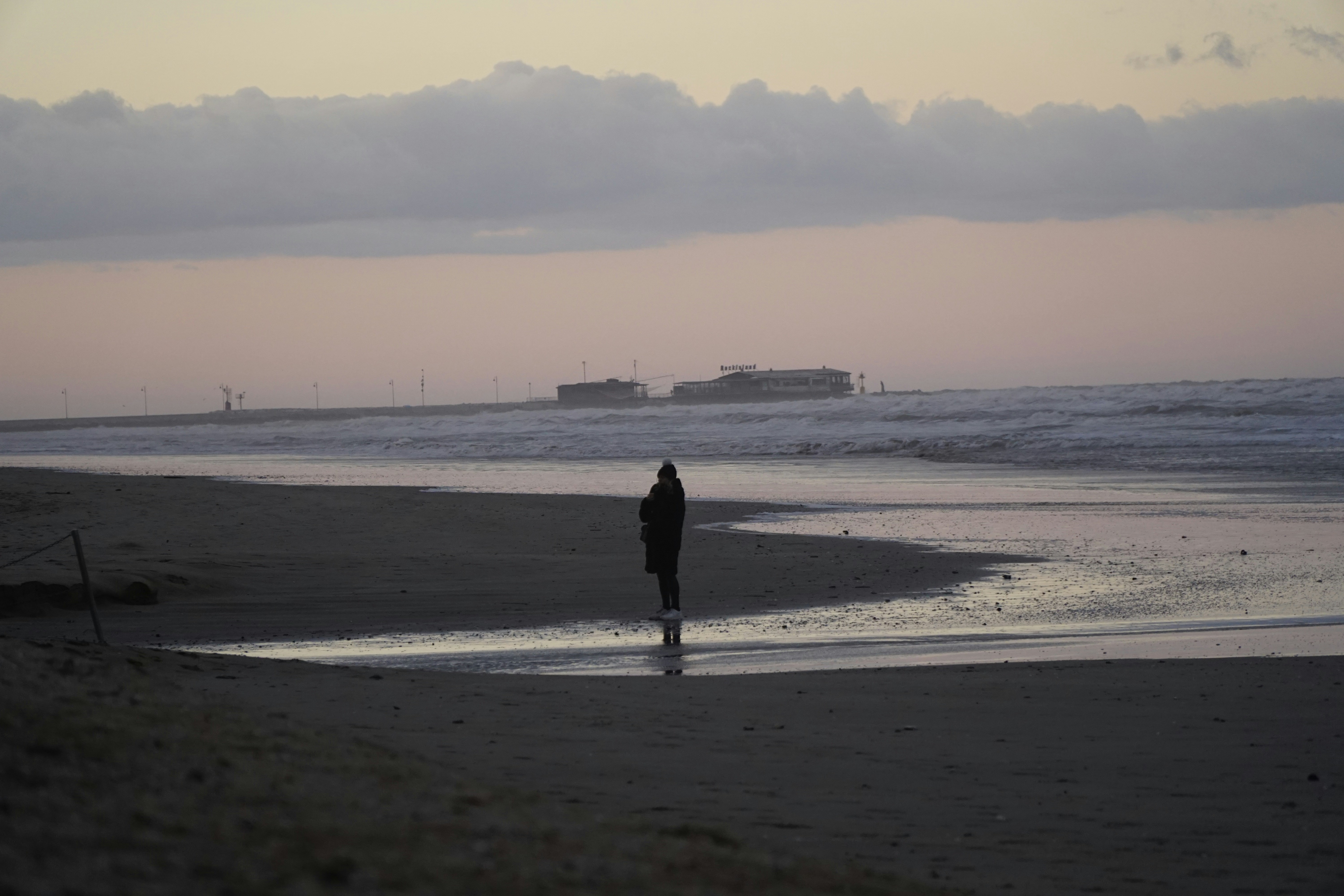 Person walking along a tranquil beach at dusk with distant islands on the horizon.