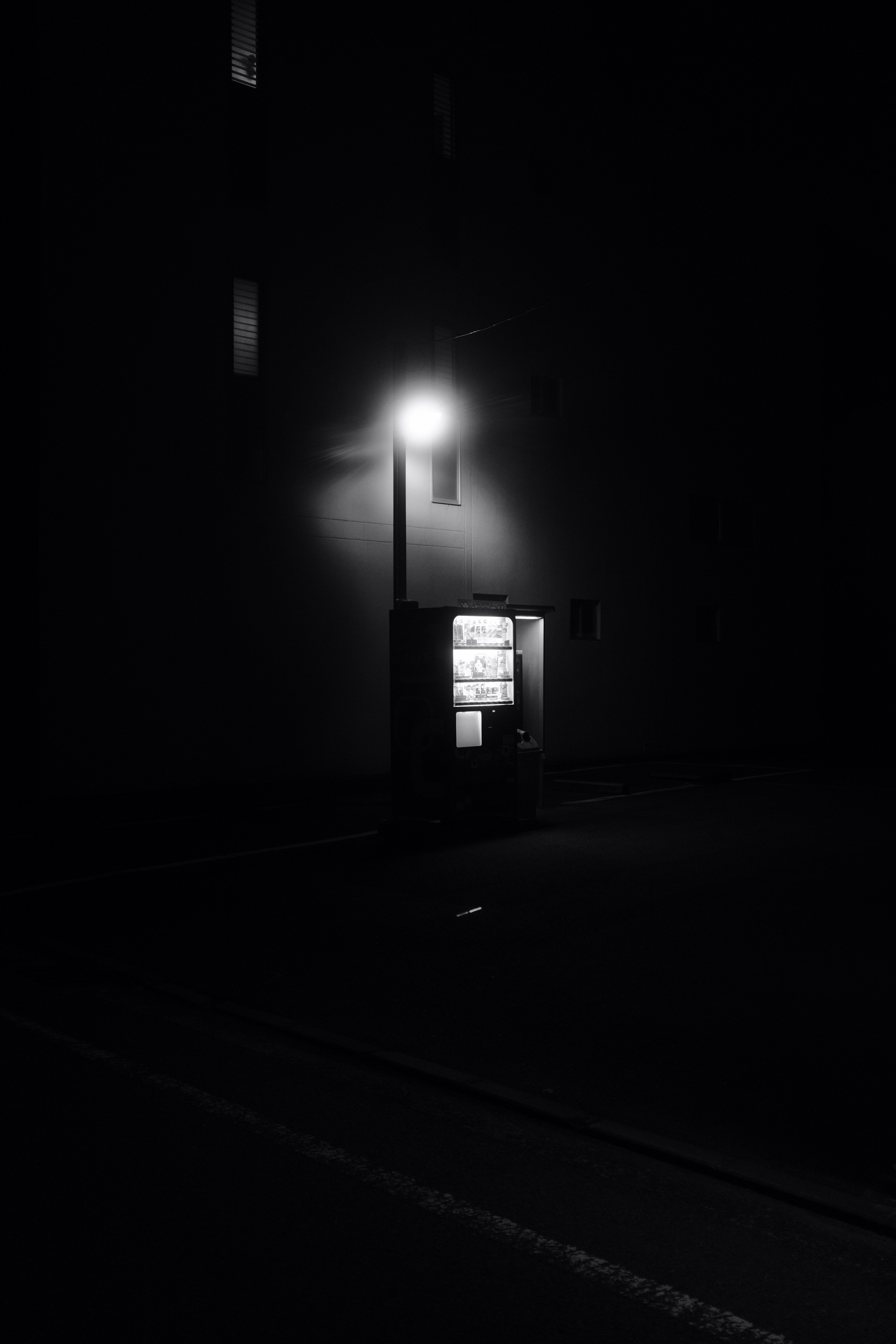 A solitary vending machine illuminated by a streetlight in a dark urban setting, creating a stark contrast against the shadows. The scene evokes a sense of isolation and intrigue.