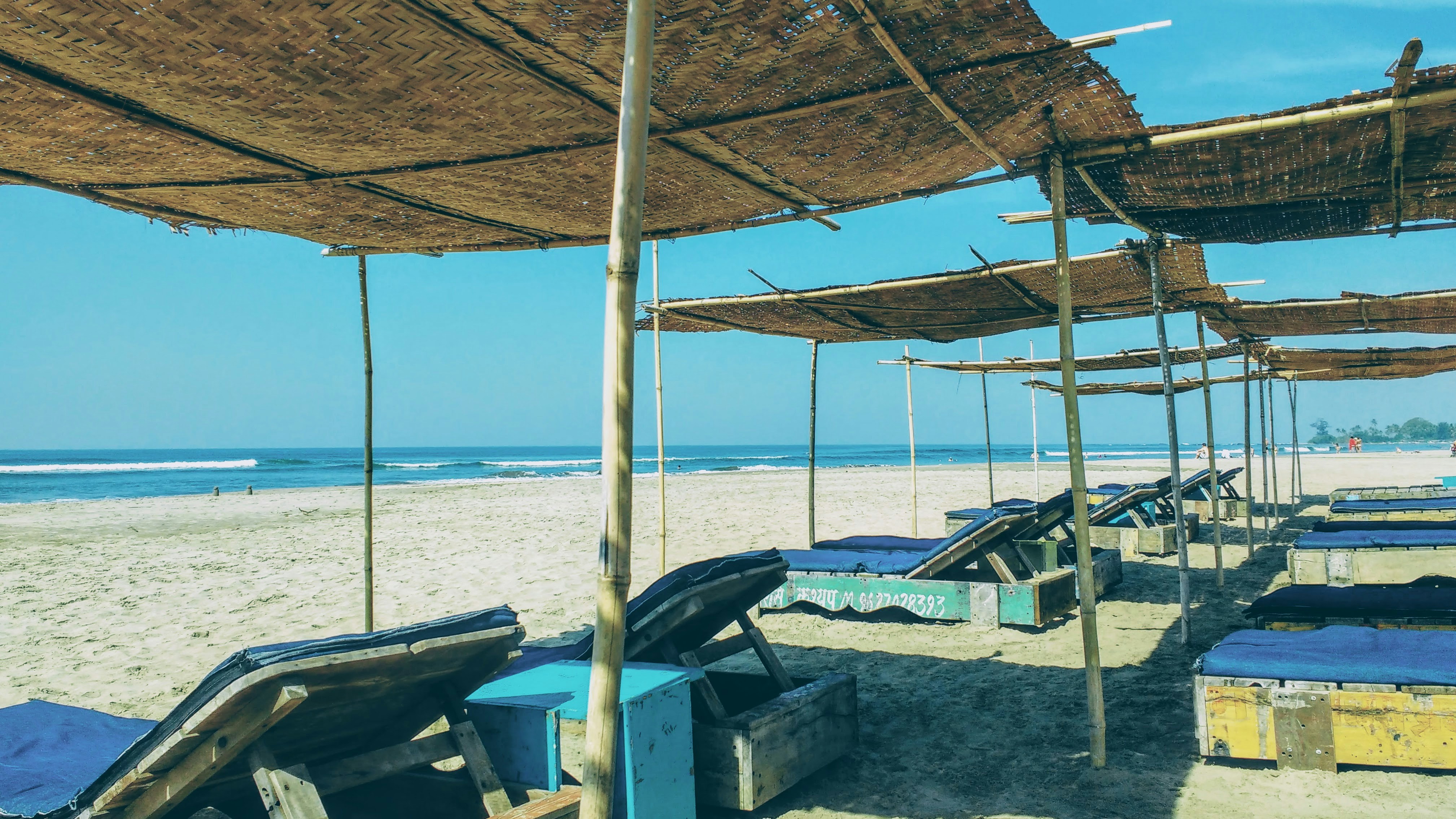 Photograph of a beach lined with straw shade canopies and lounge beds along the sand; calm sea and a clear blue sky.