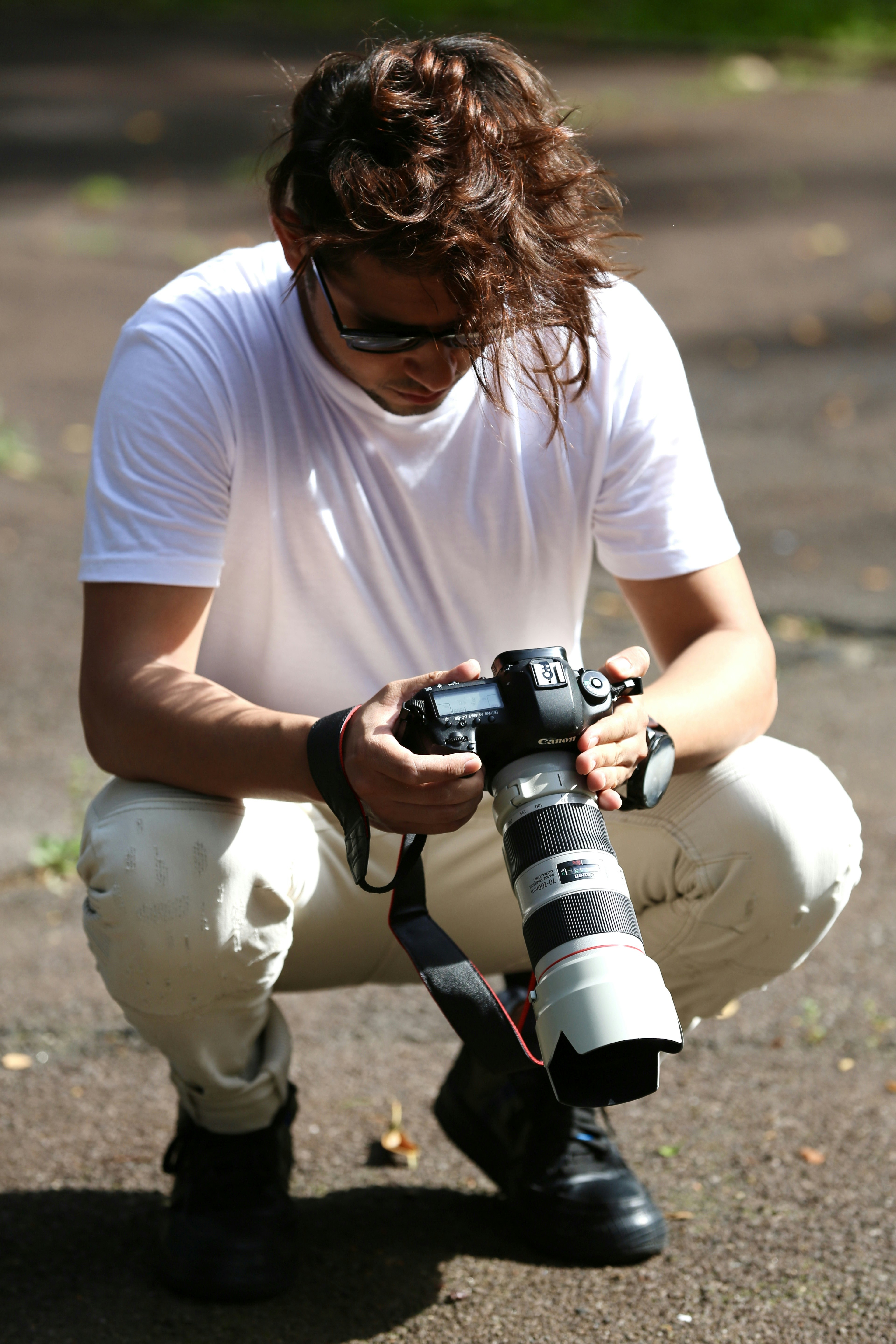 Photographer crouched down, intently examining the settings on a Canon EOS 5D Mark III with a long lens in bright sunlight.