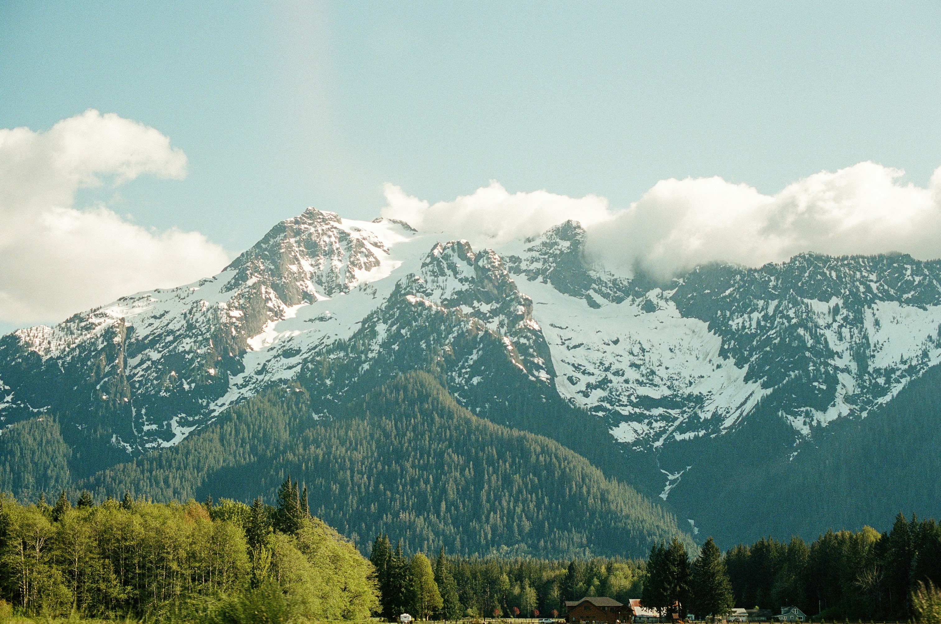 This captivating image features a majestic, snow-capped mountain range under a serene blue sky, with fluffy clouds gently wrapping around the peaks. The lush greenery at the base provides a vivid contrast to the rugged, icy summits, creating a harmonious blend of nature's elements. The soft lighting and pastel colors evoke a tranquil and awe-inspiring atmosphere, making the scene visually striking and immersive.