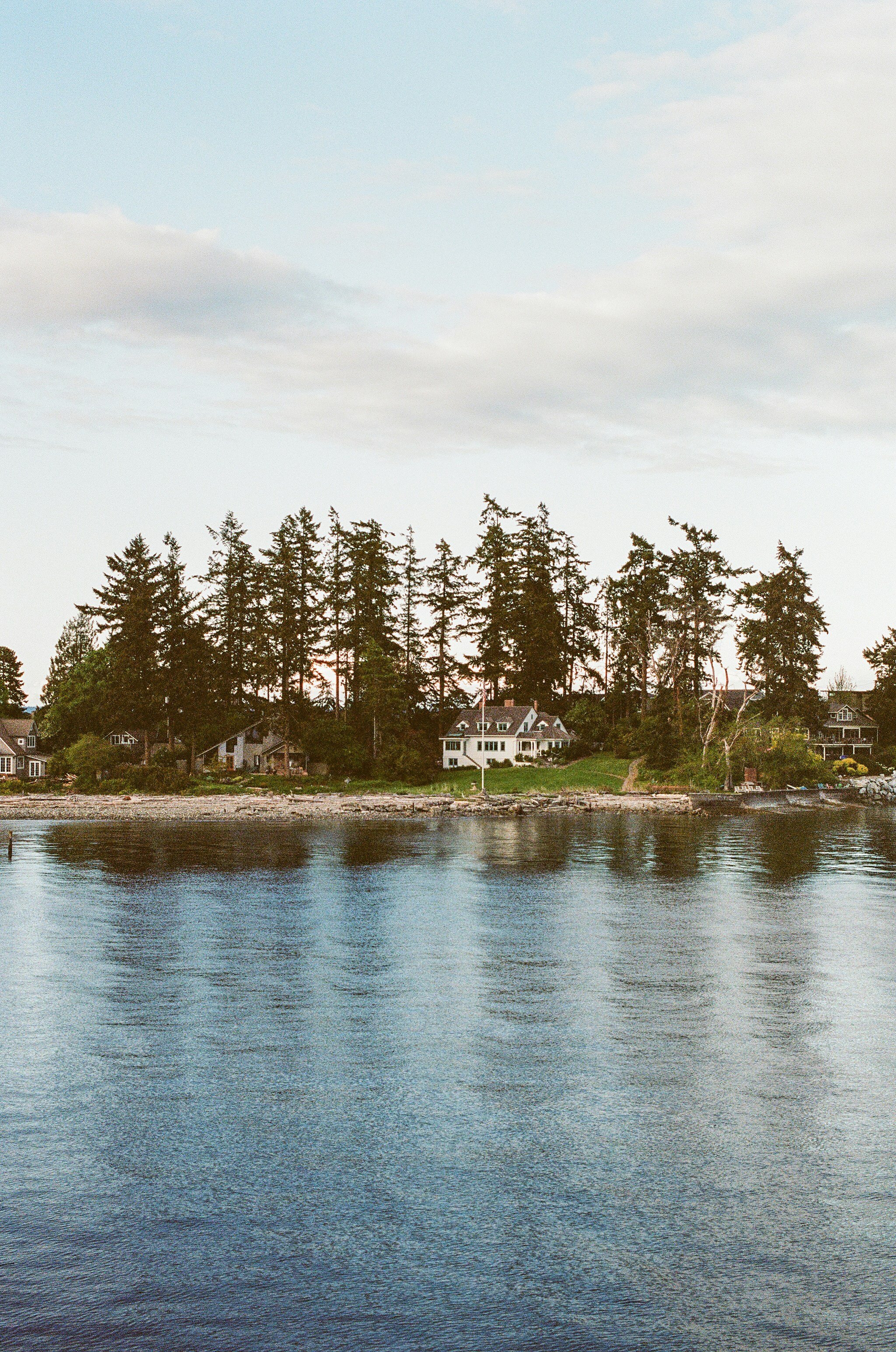 Lush trees and charming houses line a tranquil shoreline, reflecting in calm waters under a clear sky.