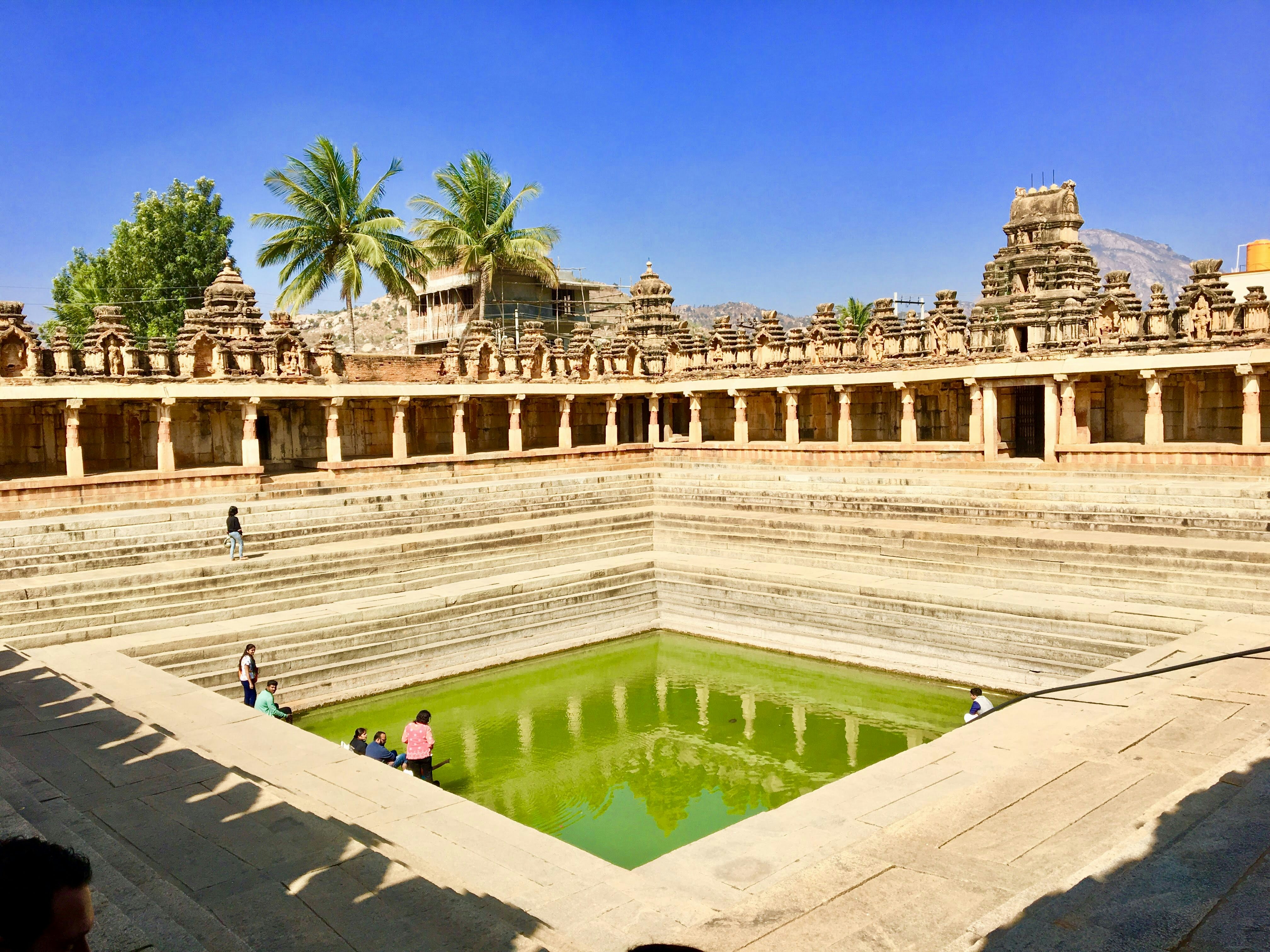 Historic stepwell surrounded by intricately carved stone walls, with a serene green pool reflecting the sky. Visitors explore the ancient structure.