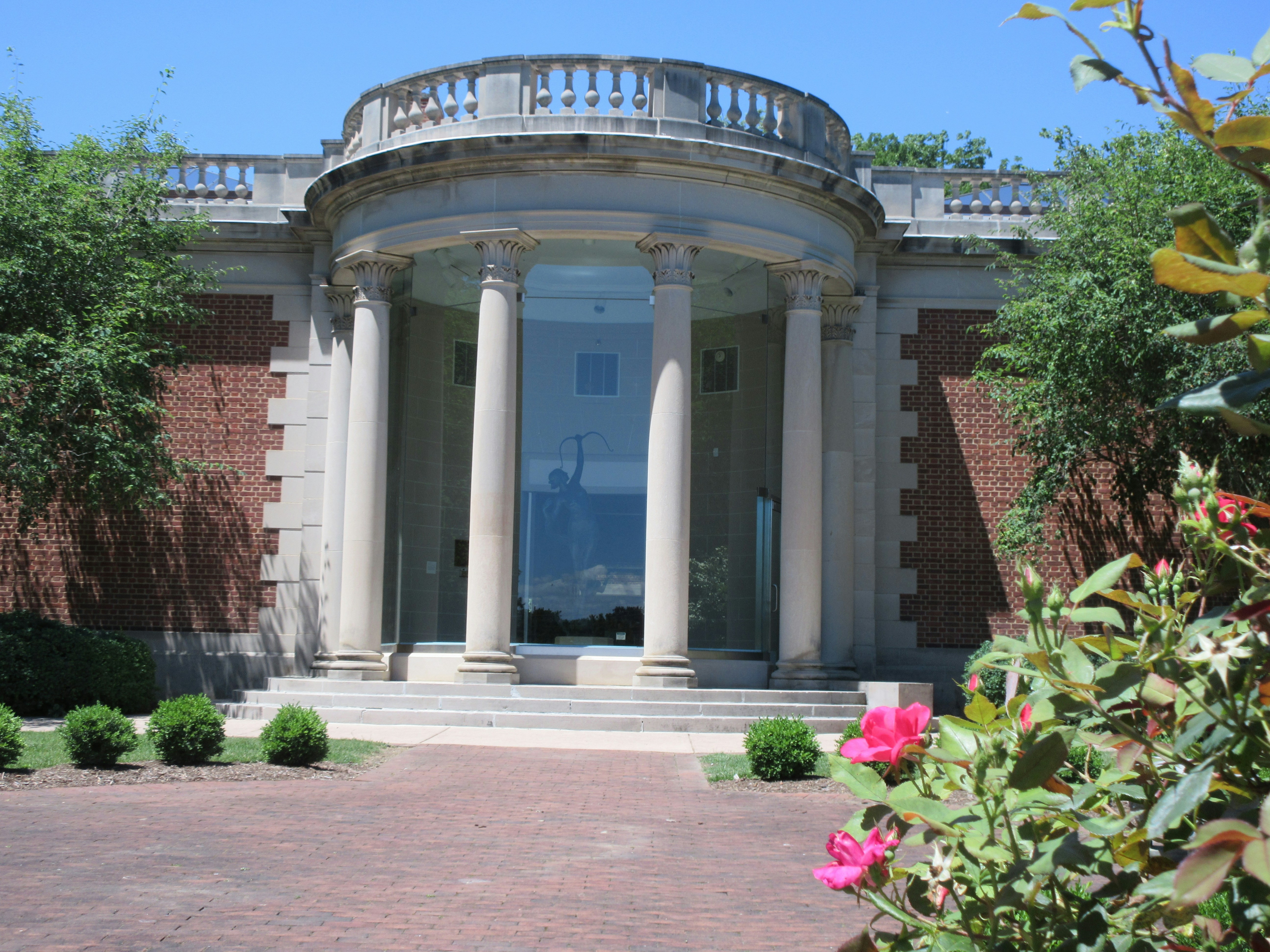 Neoclassical building entrance with tall columns and glass doors, framed by lush greenery. Pink roses bloom in the foreground, adding color contrast to the stately façade.