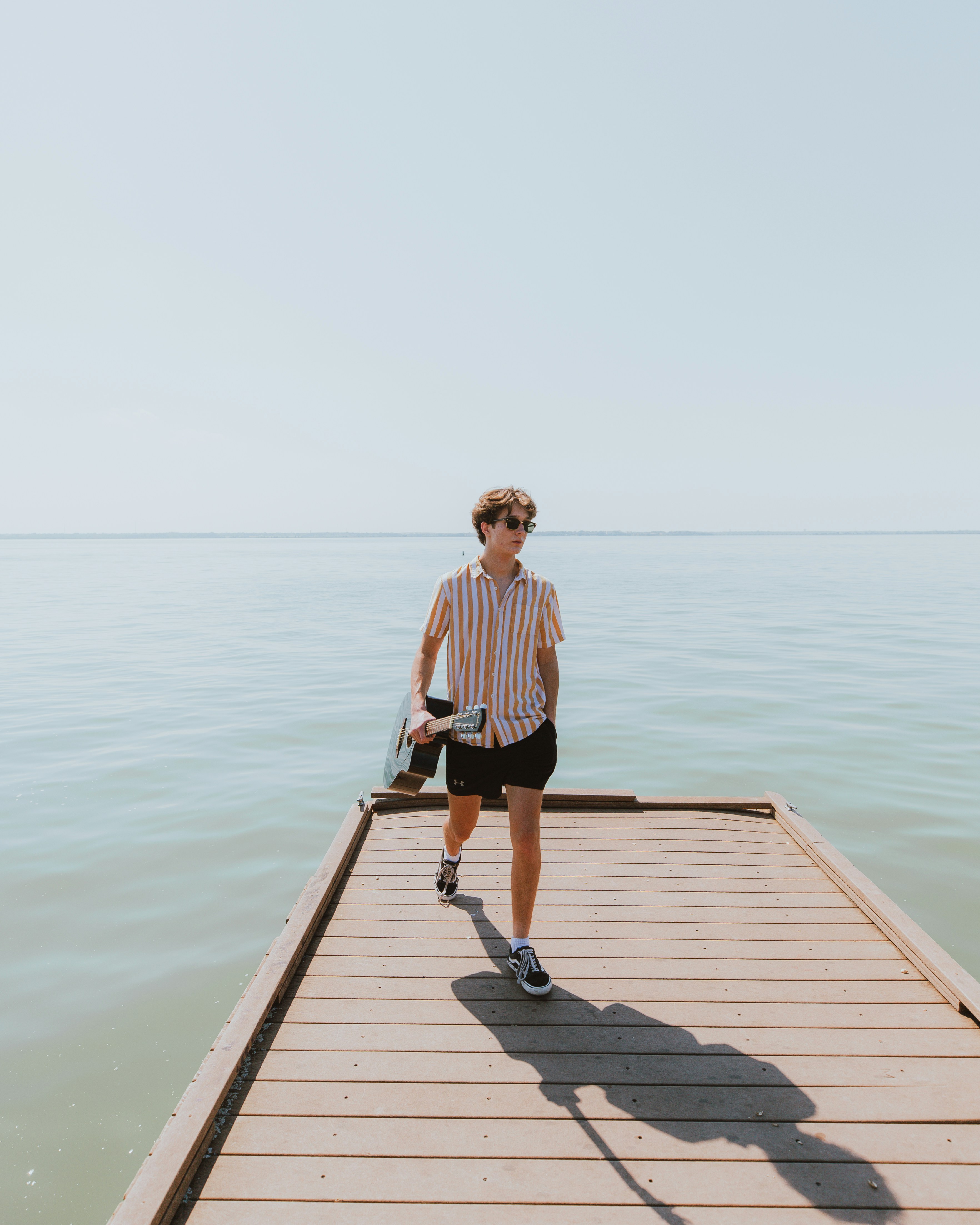Young man walking along a wooden dock by the calm waters, holding a skateboard and enjoying a sunny day.