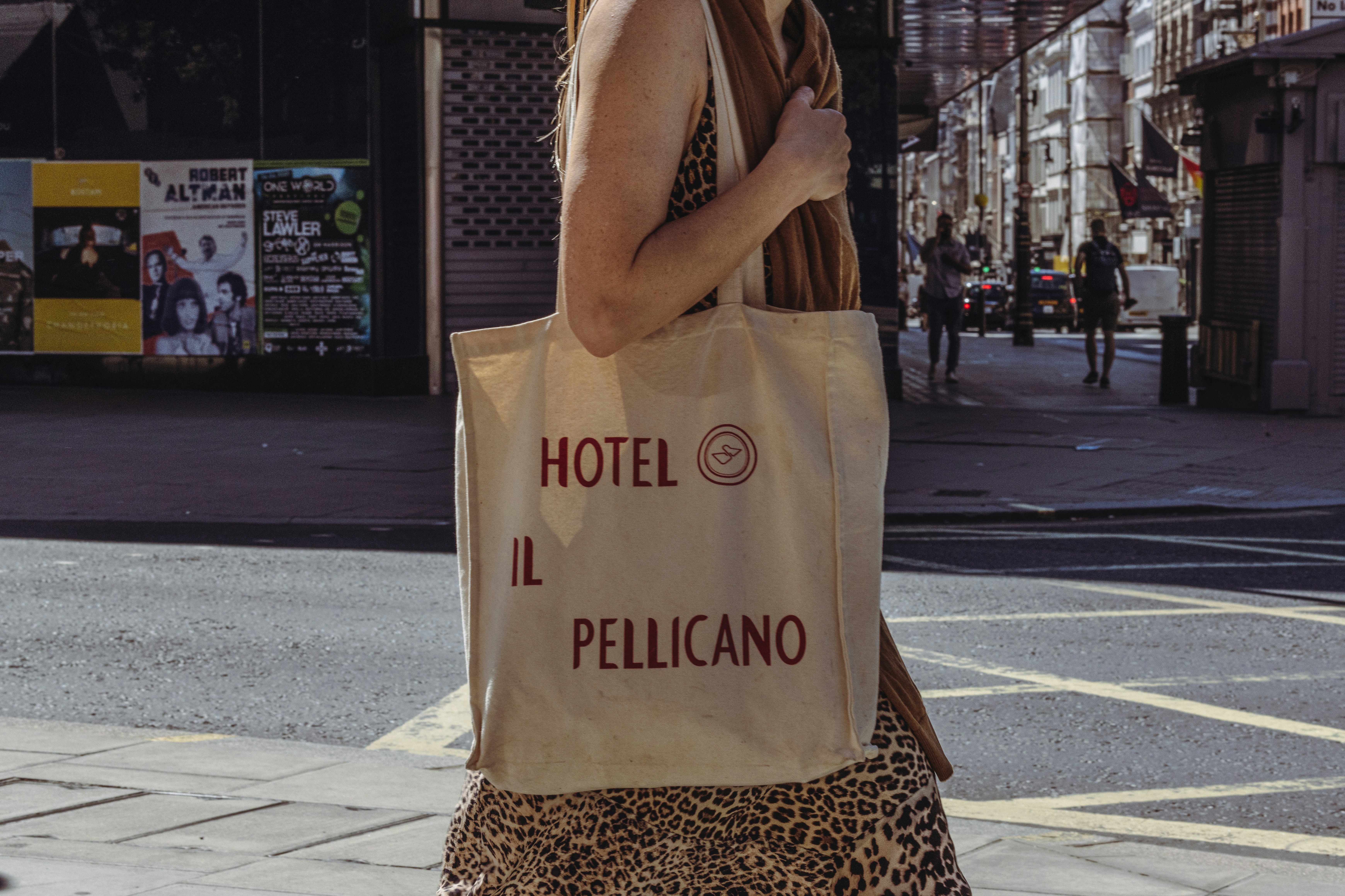 A woman walks through a bustling street, showcasing a stylish tote bag emblazoned with 'HOTEL IL PELICANO'. Her fashionable attire complements the urban backdrop.