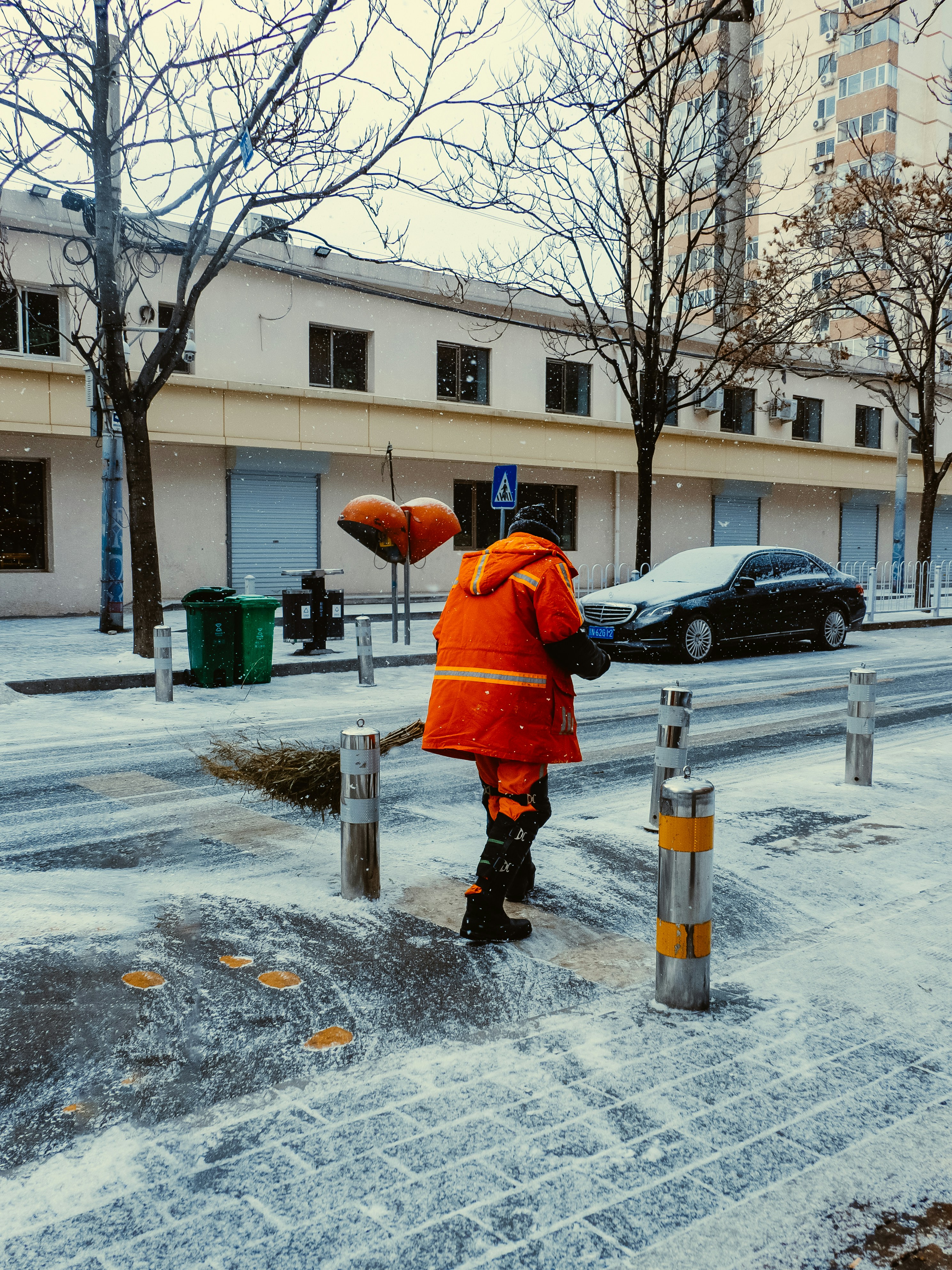 A street sweeper in an orange uniform clears snow-covered pavement amidst falling snowflakes, with urban buildings in the background.