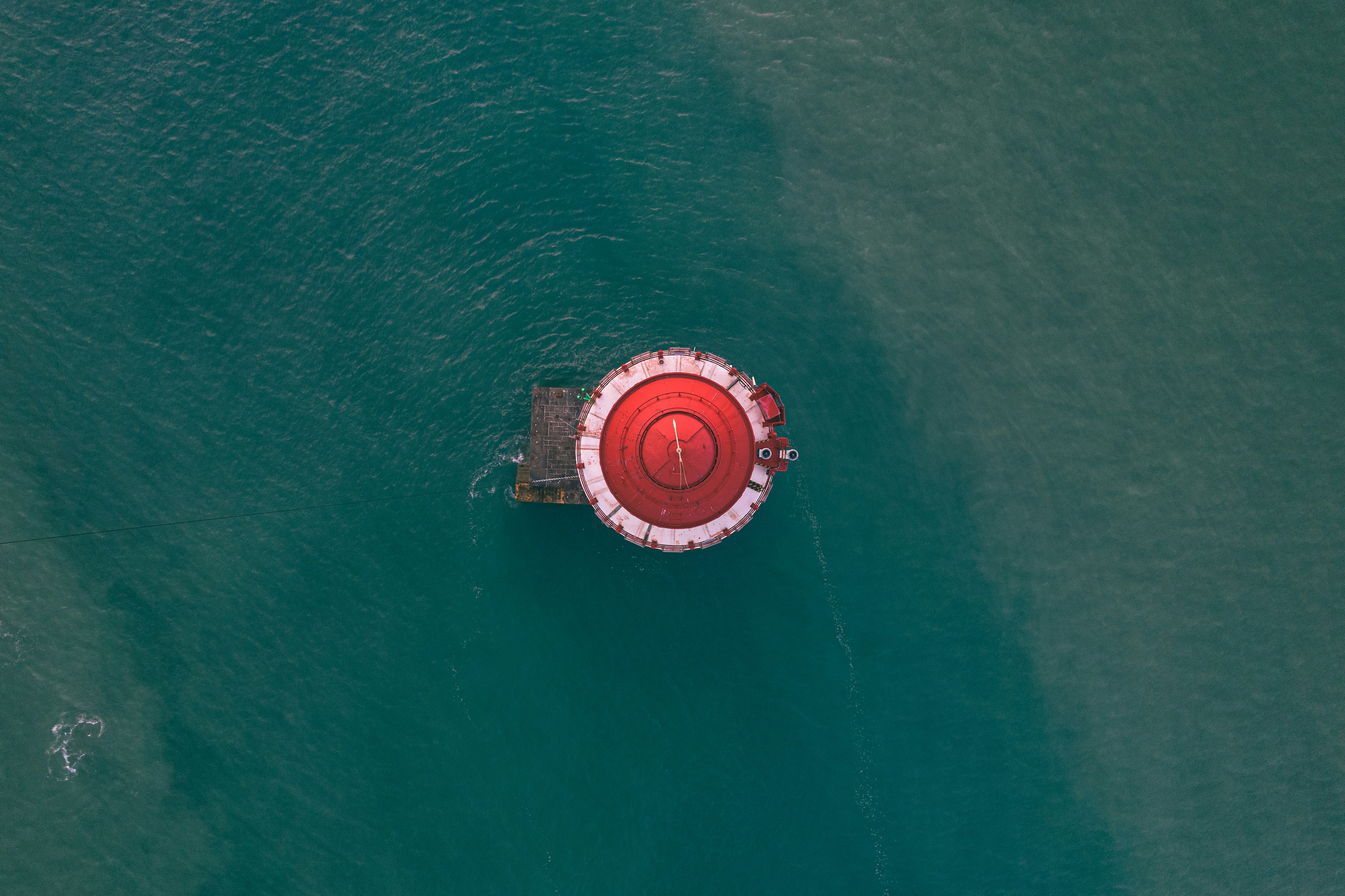 Overhead view of a lighthouse with a vibrant red roof surrounded by calm blue waters.