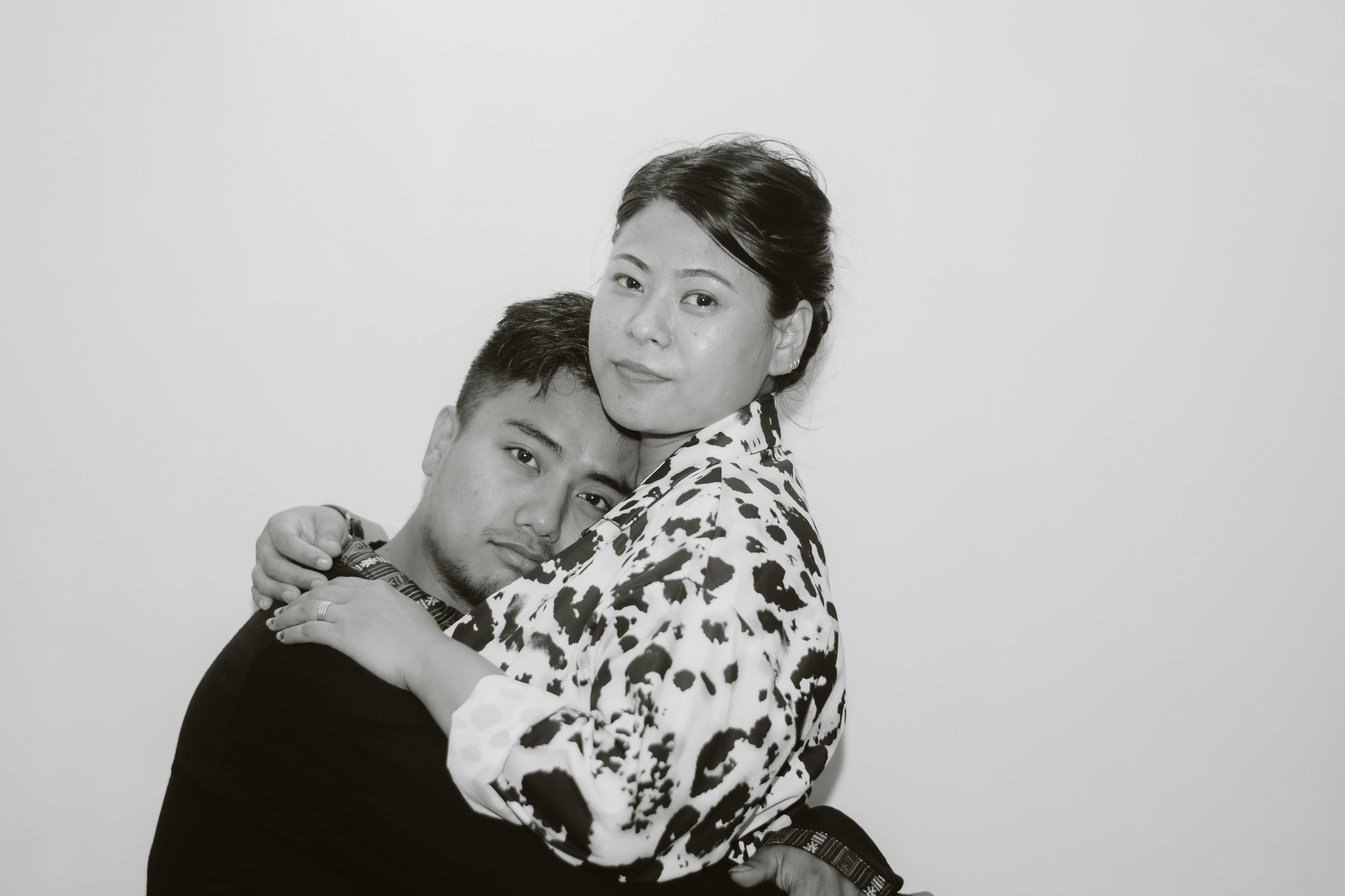 This striking monochrome photograph captures a tender moment between a couple, with one person resting their head gently on the other's shoulder. The composition is intimate, with the subjects centered against a plain background that enhances the focus on their expressions and connection. The subtle play of light and shadow creates a serene and heartfelt atmosphere, accentuating the emotional depth of the scene.