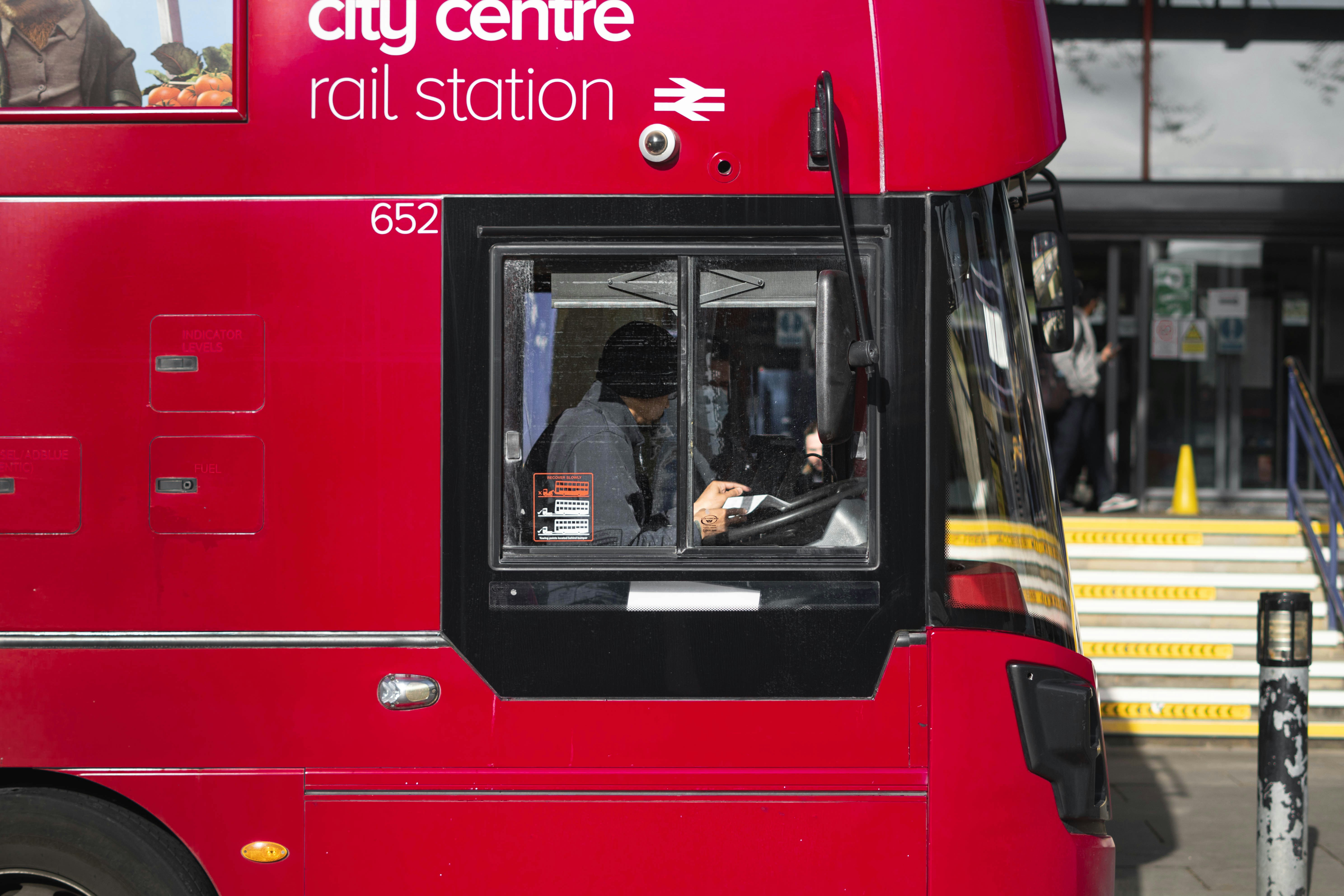 Red double-decker bus parked at a city rail station, with a driver preparing for departure.