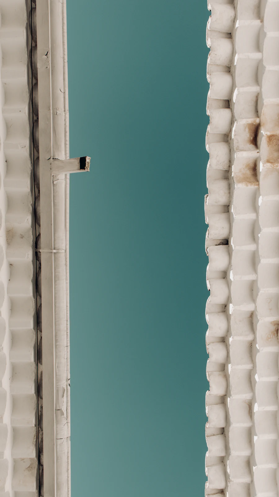 A vertical slice of teal sky framed by two pale, textured building edges and a small protruding bracket on the left. The composition feels abstract and architectural.