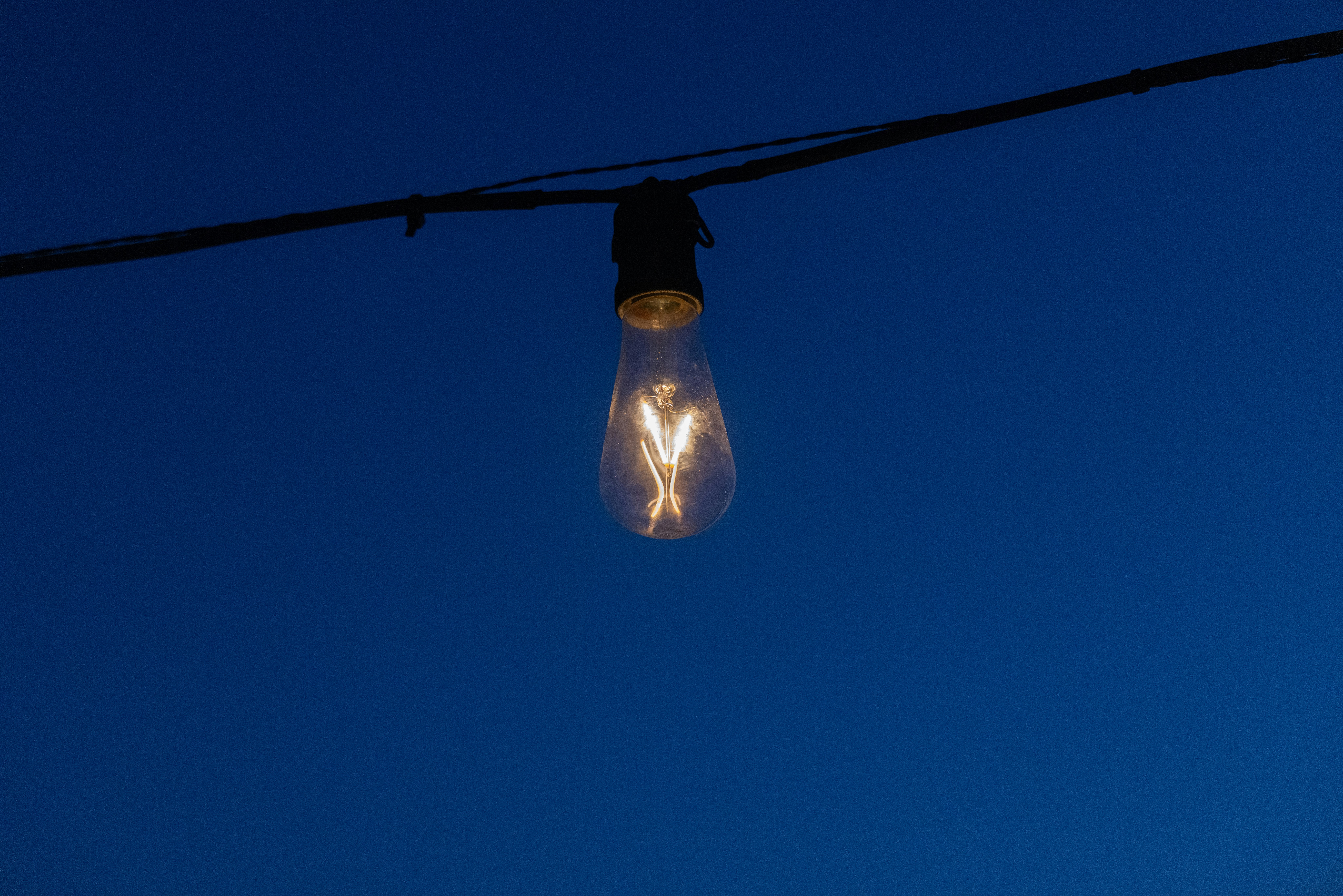 A vintage light bulb glowing against a deep blue twilight sky, suspended from a wire. The intricate filament design adds a nostalgic touch.