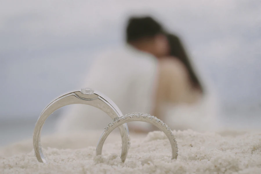 Two wedding rings resting in sand with a softly blurred couple in the background by the water. Gentle pastel tones create a romantic seaside scene.