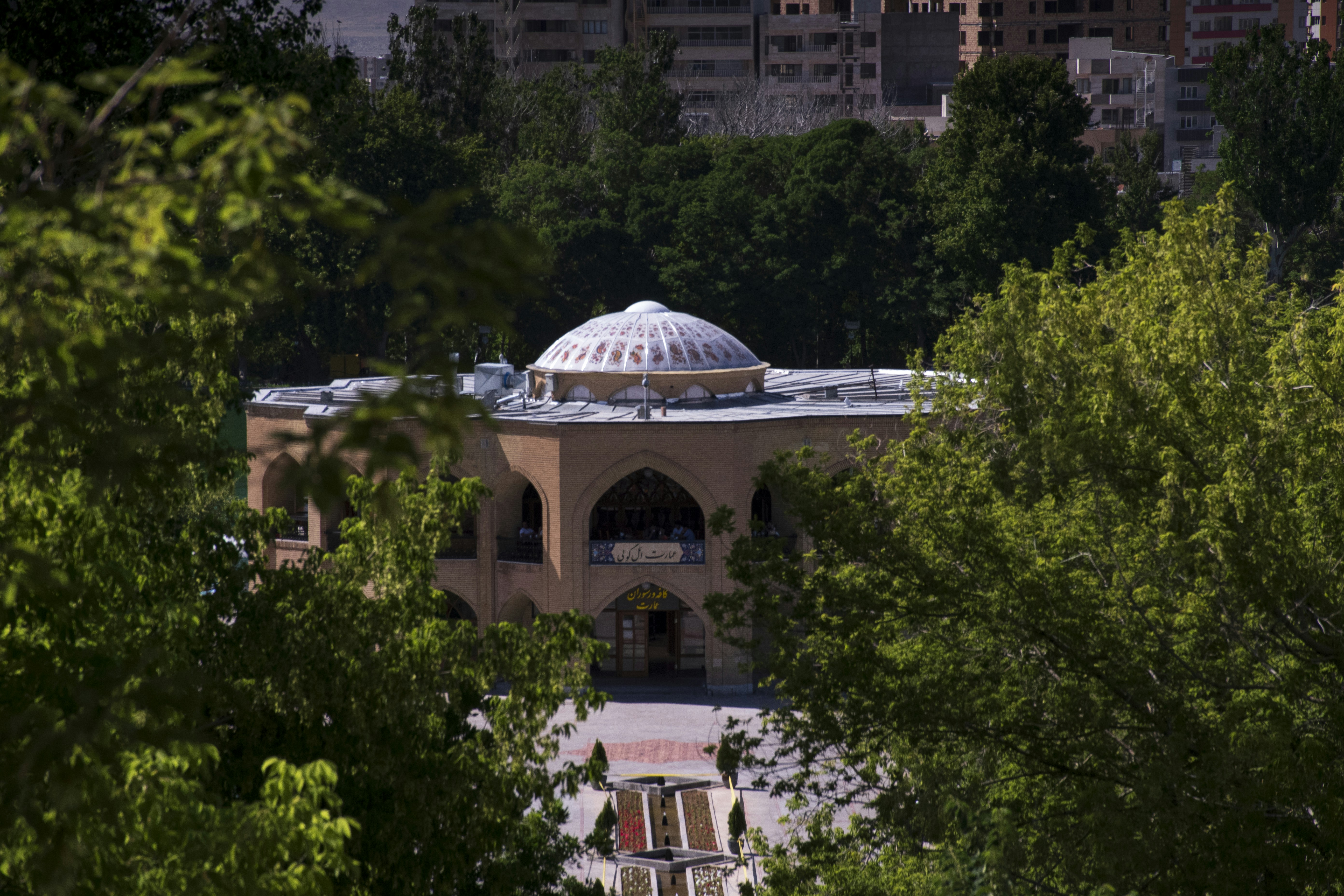 Ornate building with a domed roof surrounded by lush trees and urban high-rises.