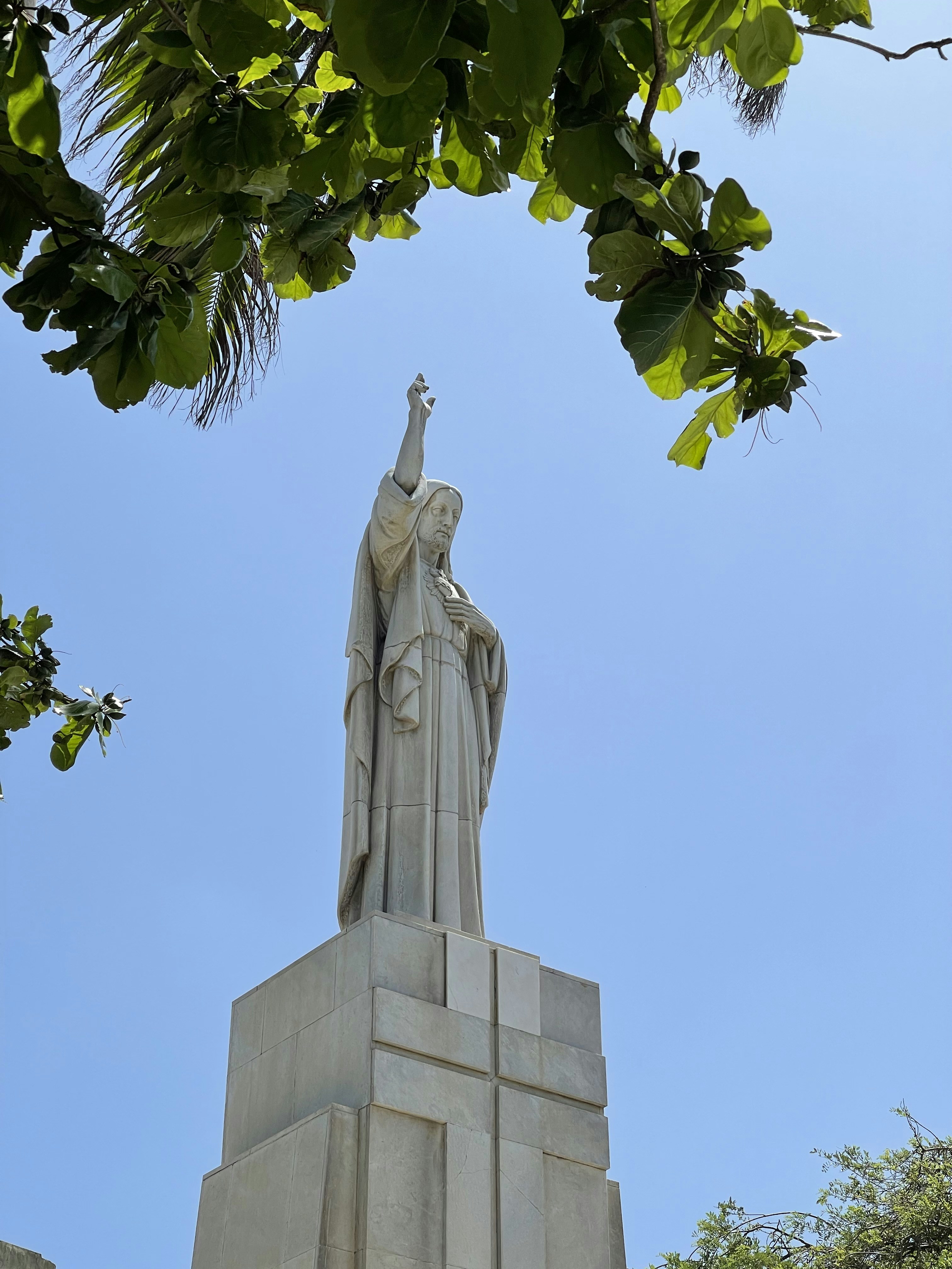 Statue of a figure with outstretched arm, framed by lush green leaves under a clear blue sky.