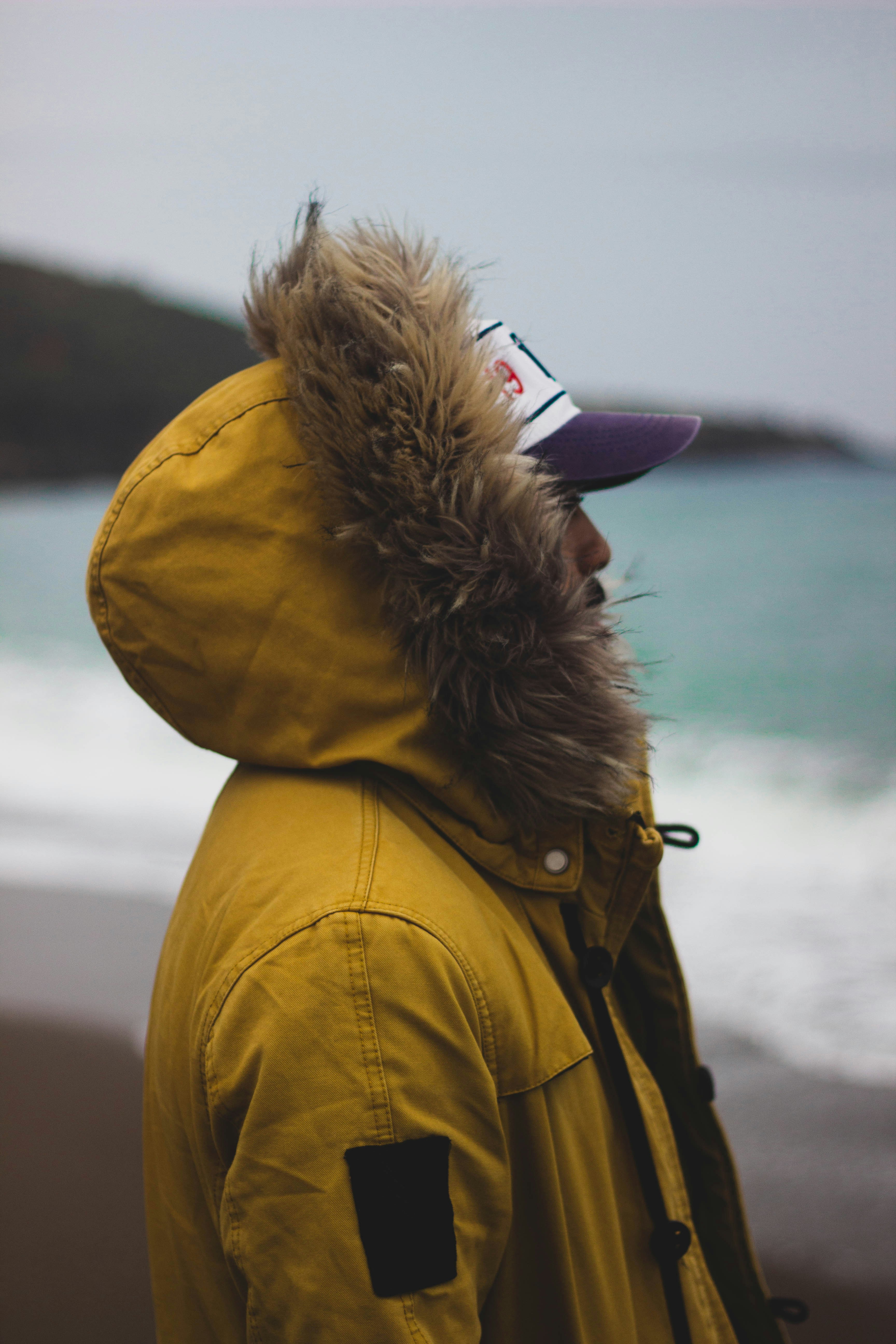 A figure in a yellow parka stands contemplatively on a beach, gazing at the ocean waves under a cloudy sky.