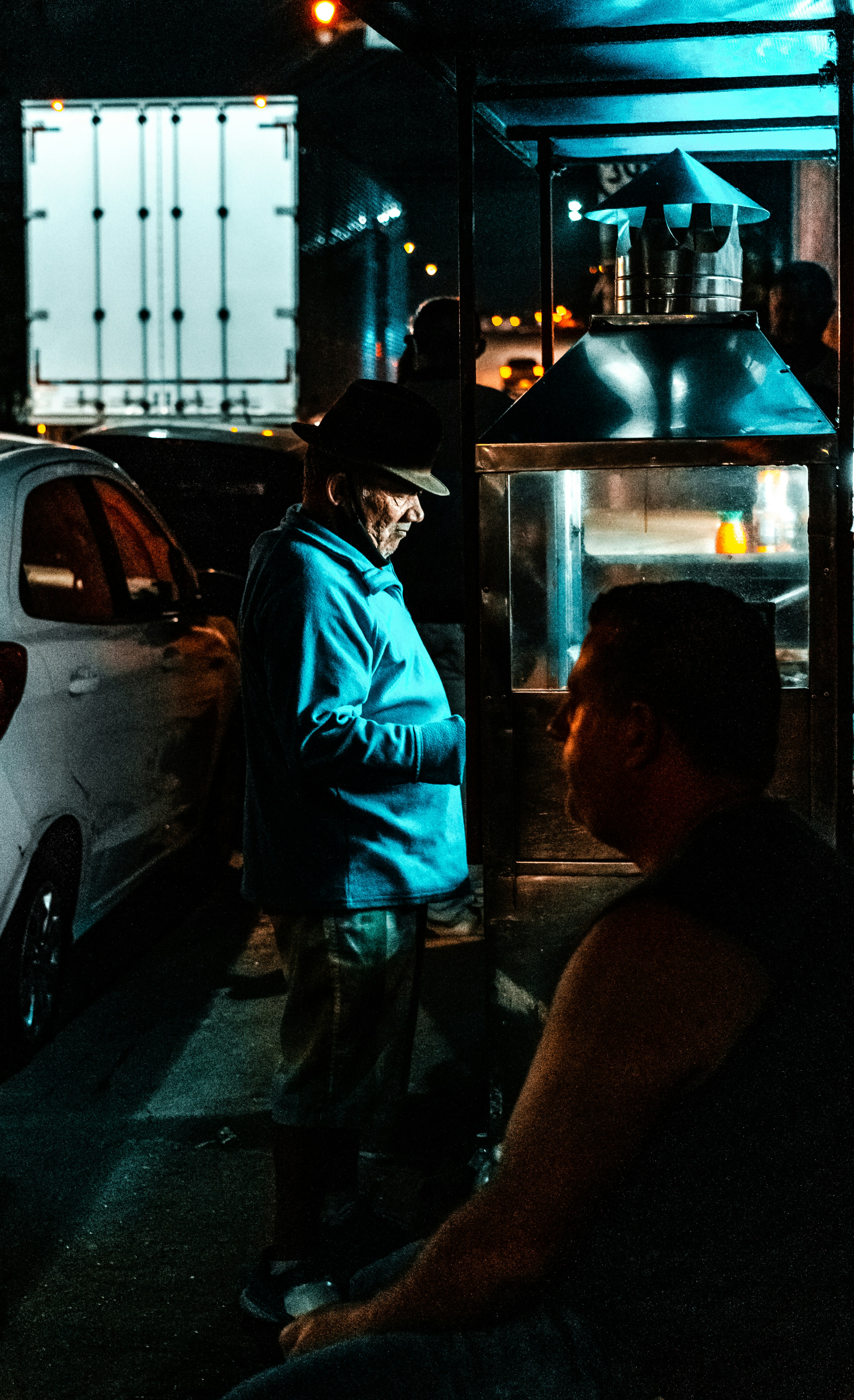 An elderly man in a blue jacket stands pensively by a street food stand, illuminated by soft light, while a blurred figure sits nearby. The urban night scene captures a moment of contemplation amid bustling city life.