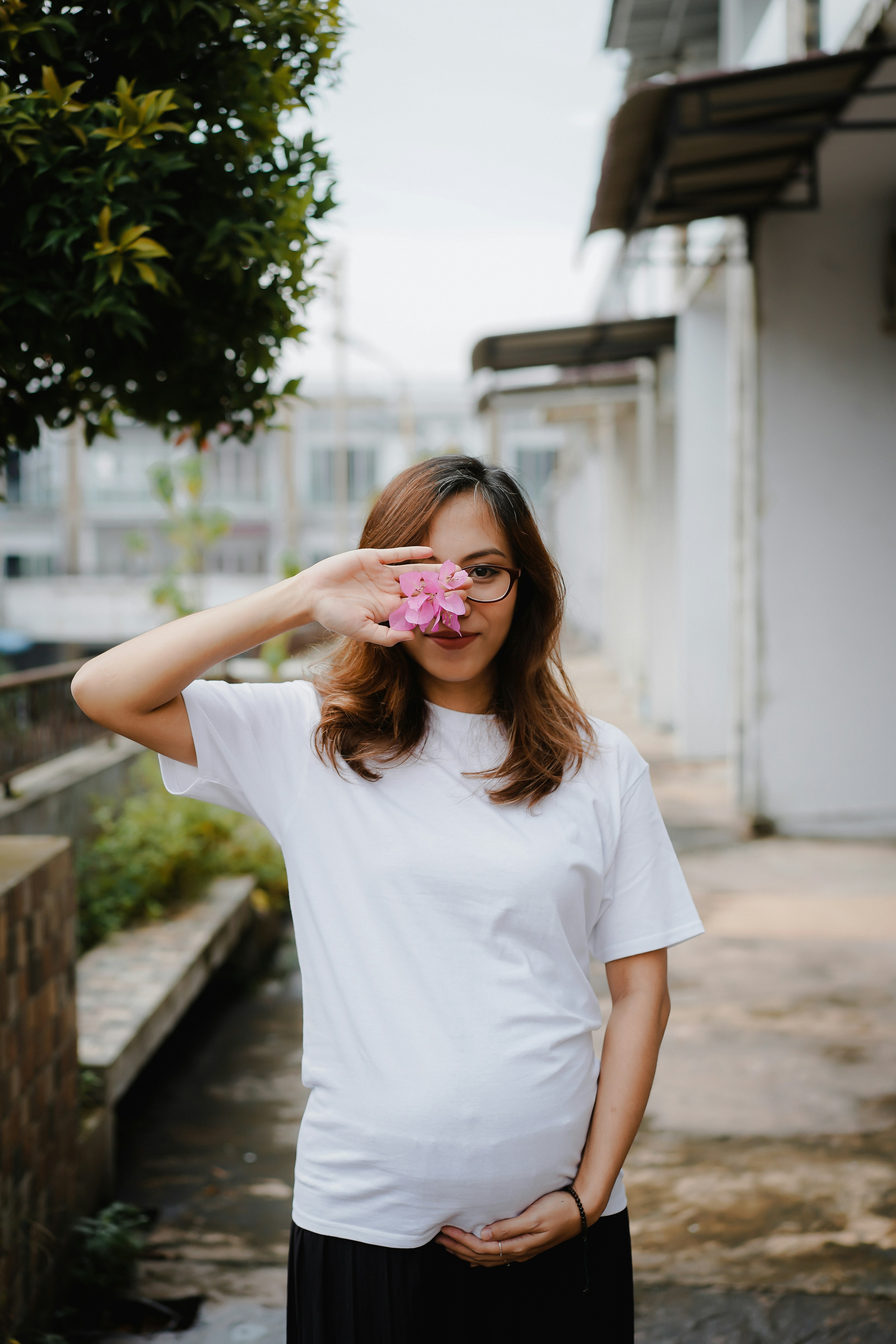 Pregnant woman holding a pink flower in front of her face, standing in a serene outdoor setting. The soft light enhances the tranquil atmosphere.