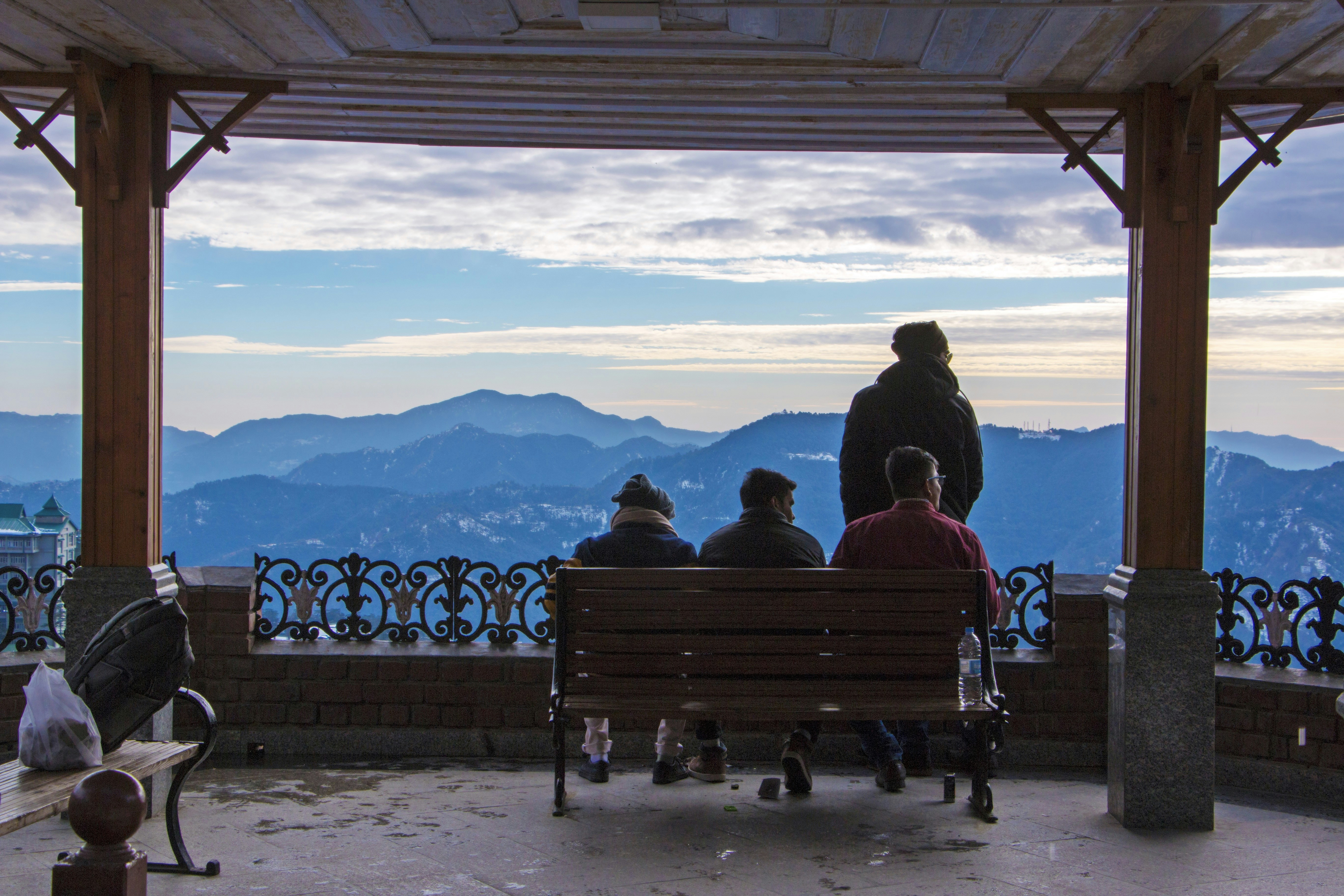 Three people seated on a bench under a pavilion, gazing at distant mountains and a vibrant morning sky.