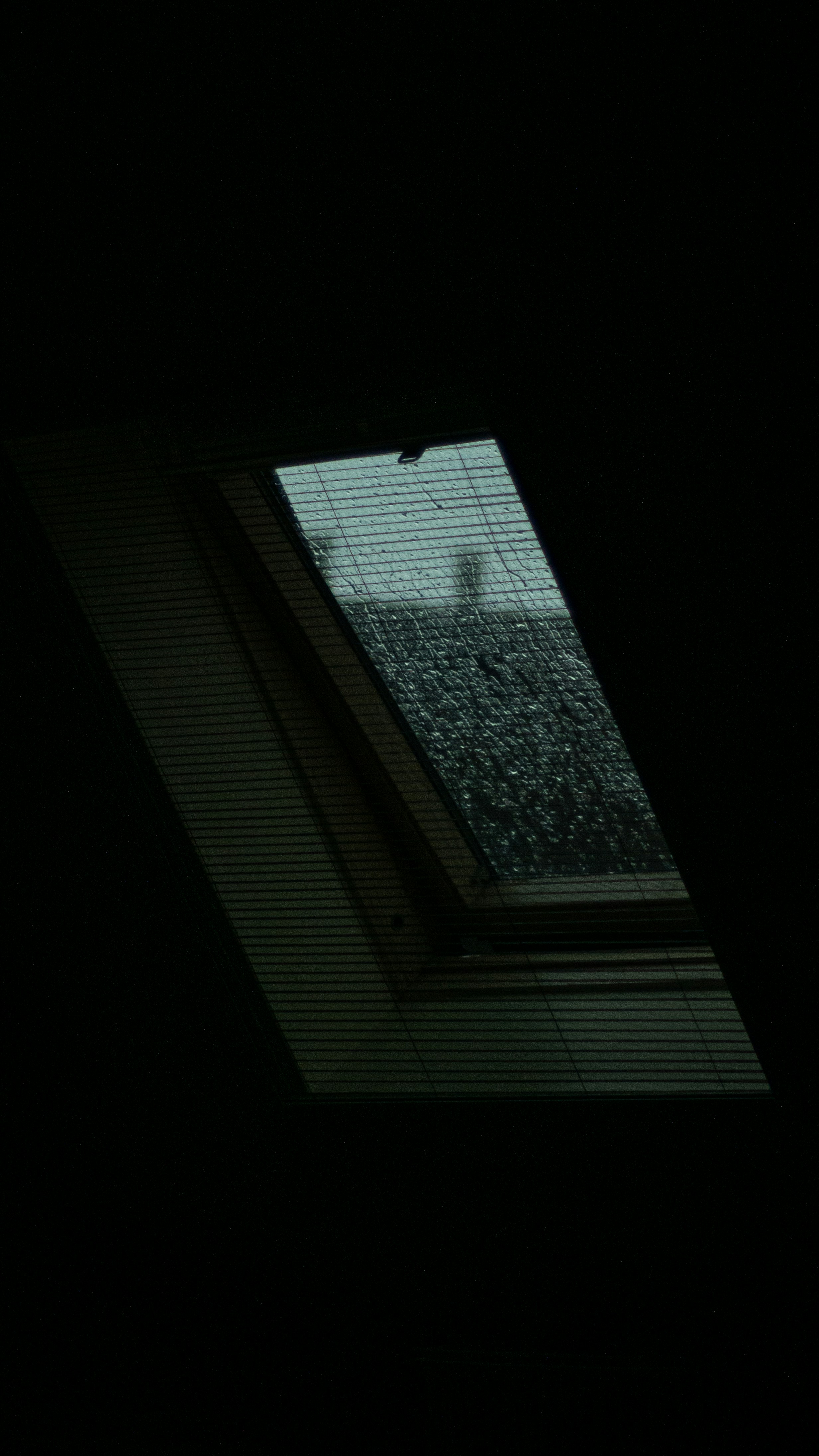 Raindrops cascading down a skylight, framed by the shadows of a dimly lit room. The blinds partially obscure the view outside.