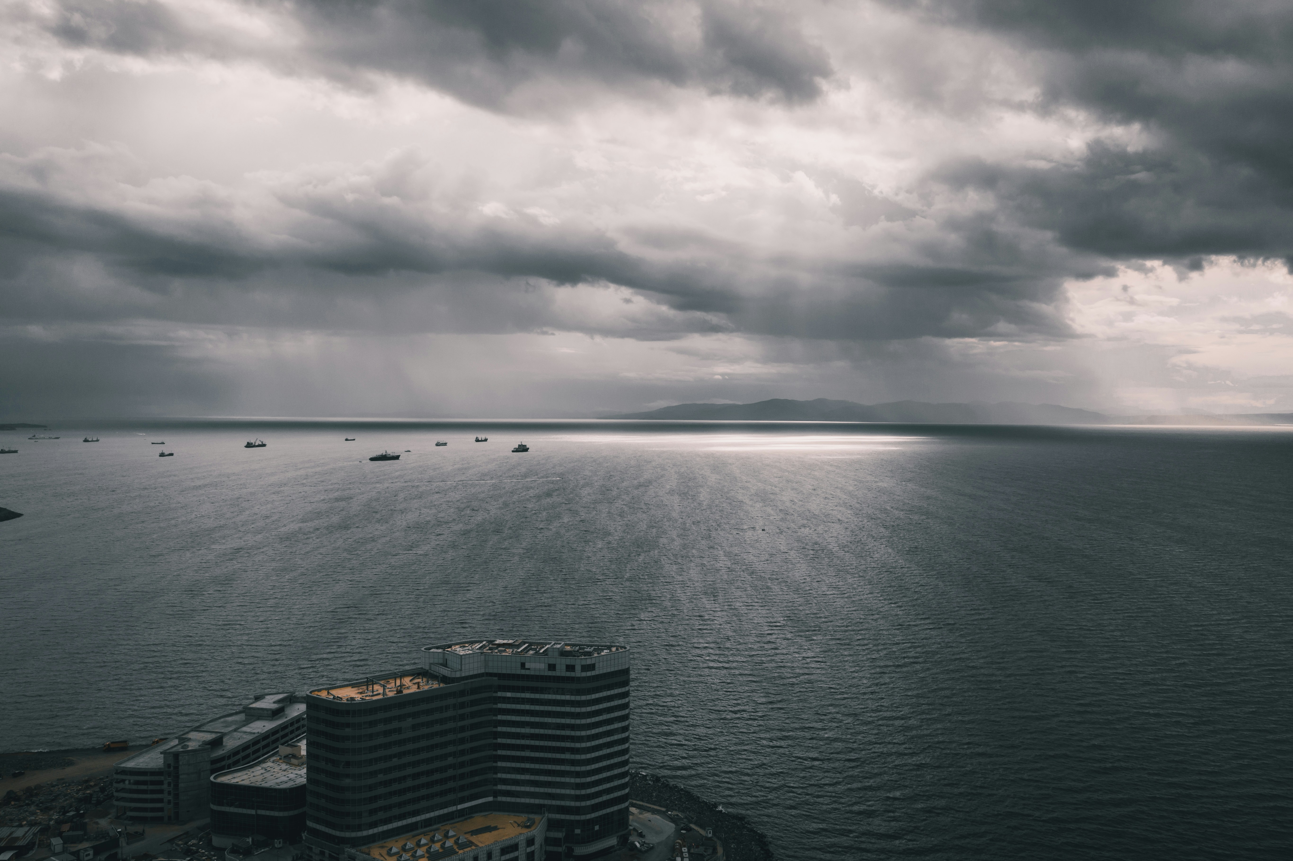 A moody seascape featuring a distant island under dramatic clouds, with sunlight breaking through, illuminating the water's surface. The foreground showcases a modern building overlooking the ocean.